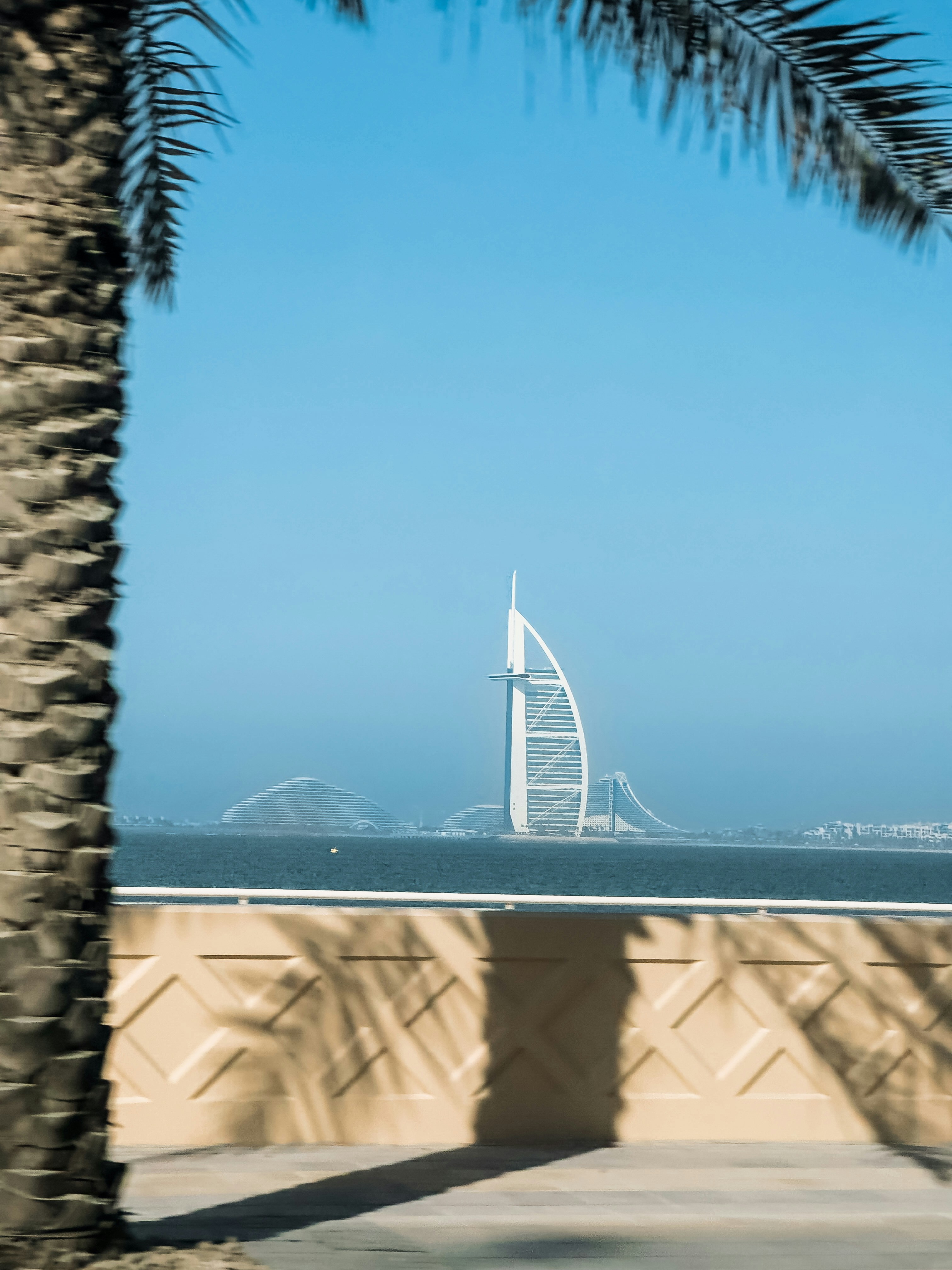Iconic Burj Al Arab rising above the serene waters, framed by palm leaves and a sandy foreground.