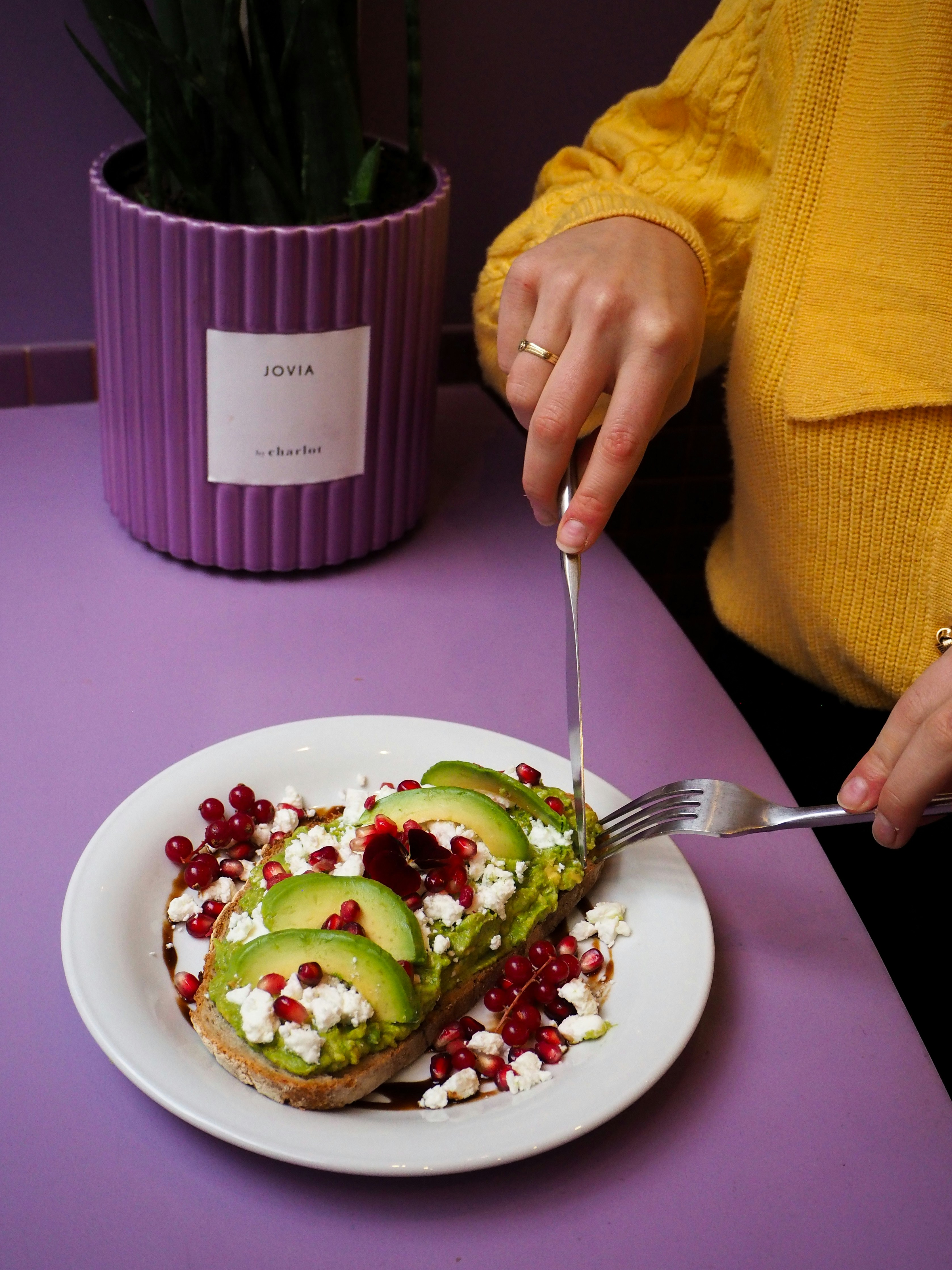 Avocado toast topped with fresh slices of apple and pomegranate seeds, elegantly presented on a white plate. A hand prepares to slice through the delicious dish.