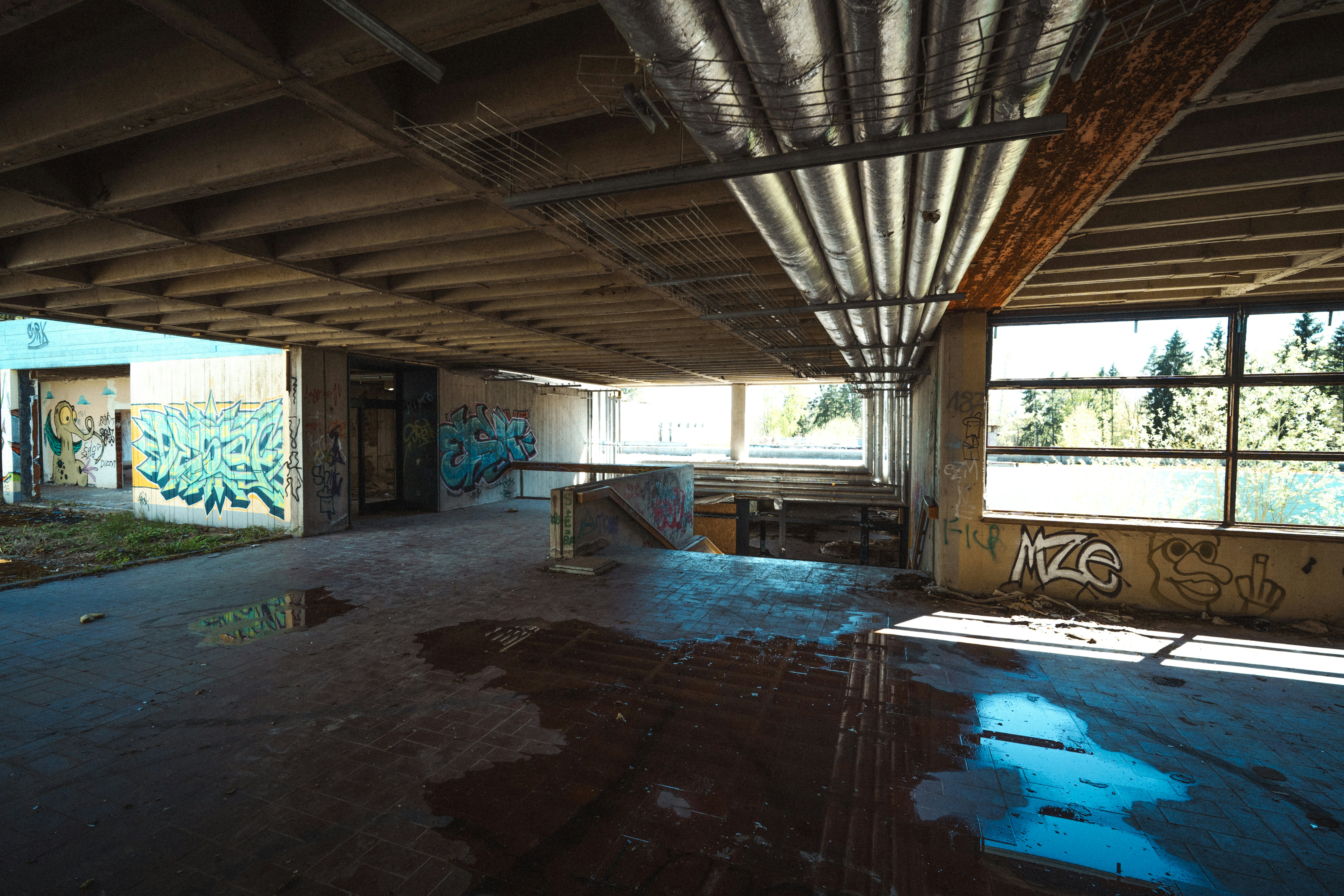 Abandoned interior space featuring graffiti on walls and water pooling on the floor, illuminated by natural light streaming through large windows.