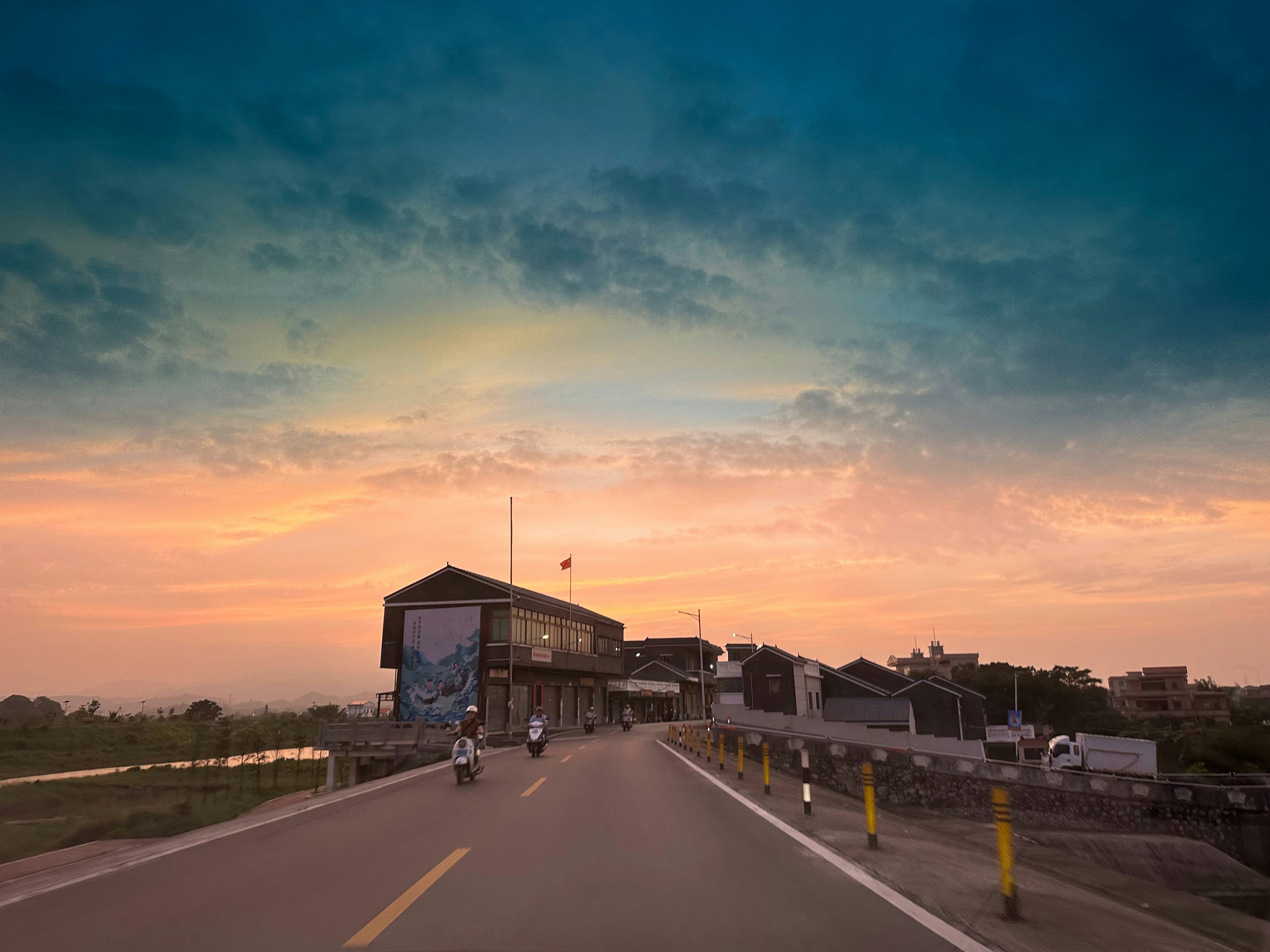 Road leading to a modern building against a vibrant sunset sky, with motorbikes in motion. The scene captures the tranquility of evening travel.