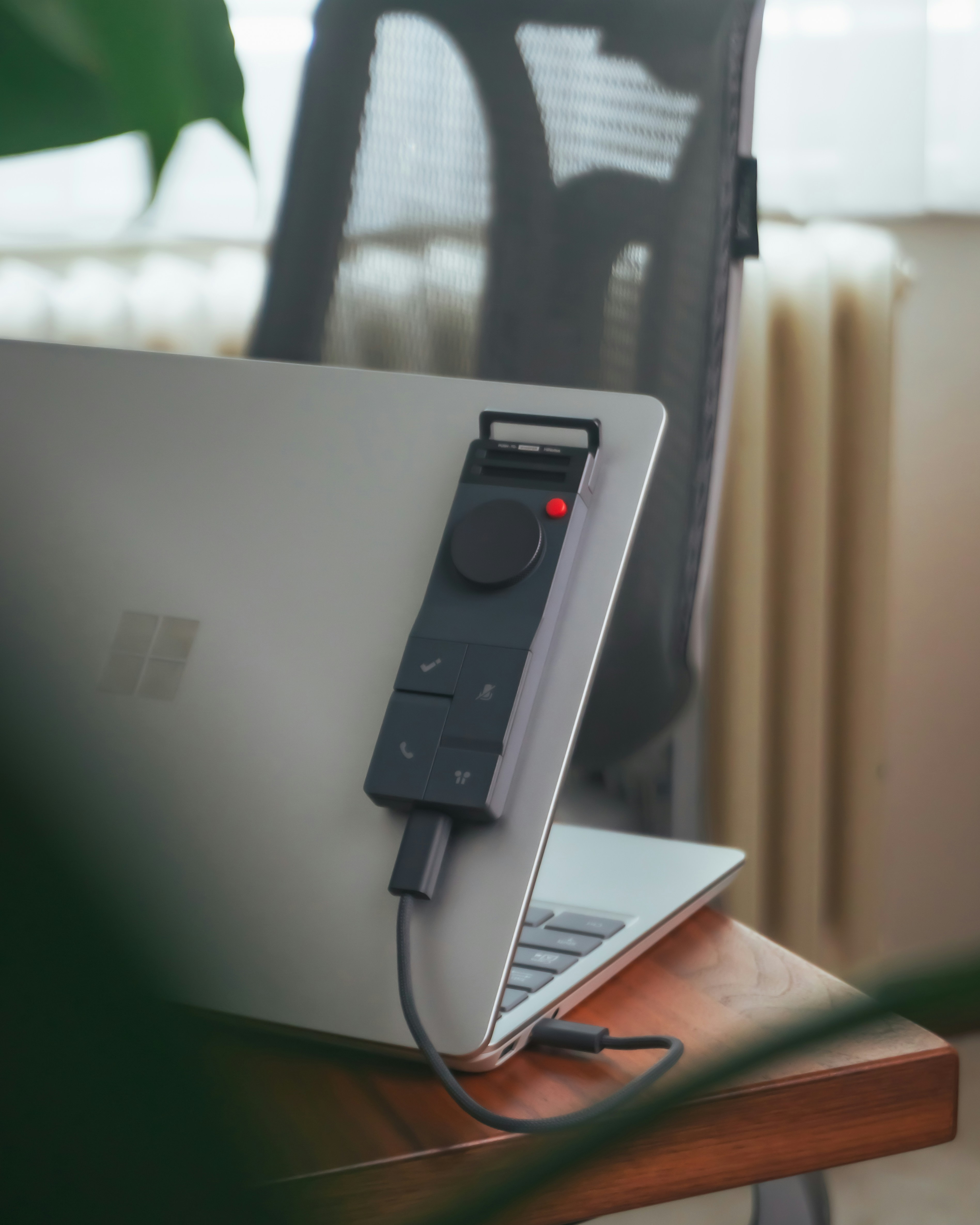 A sleek device attached to a laptop, featuring buttons for calls and a prominent red recording light. The scene is set in a contemporary workspace with a hint of greenery.