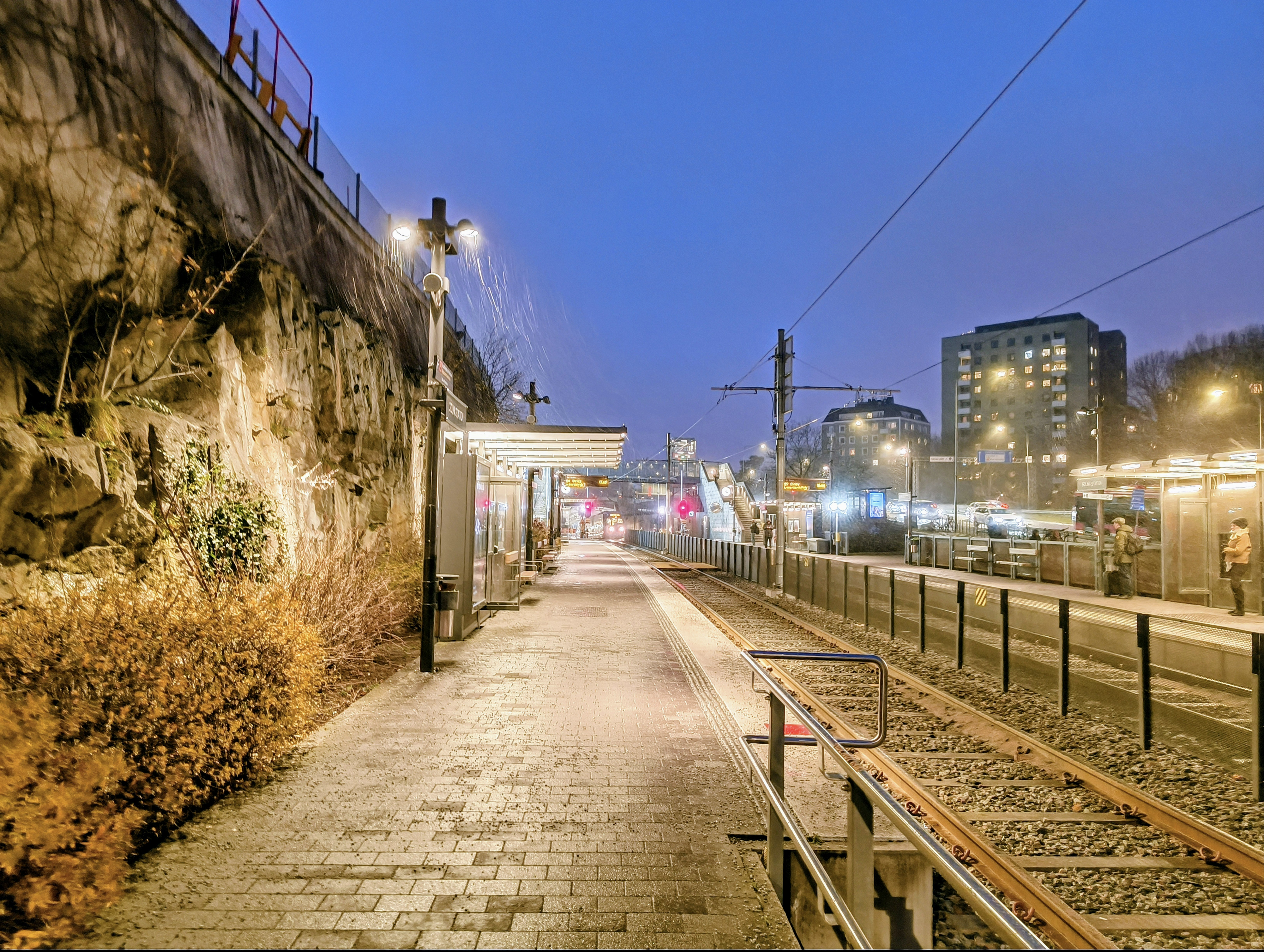 A tranquil scene of a deserted train station with illuminated pathways and a backdrop of urban buildings under a twilight sky.