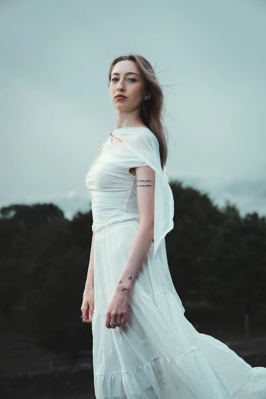 A woman in a flowing white dress stands outdoors against a pale sky and dark tree line. Her face is turned toward the camera, with the fabric trailing in the wind.