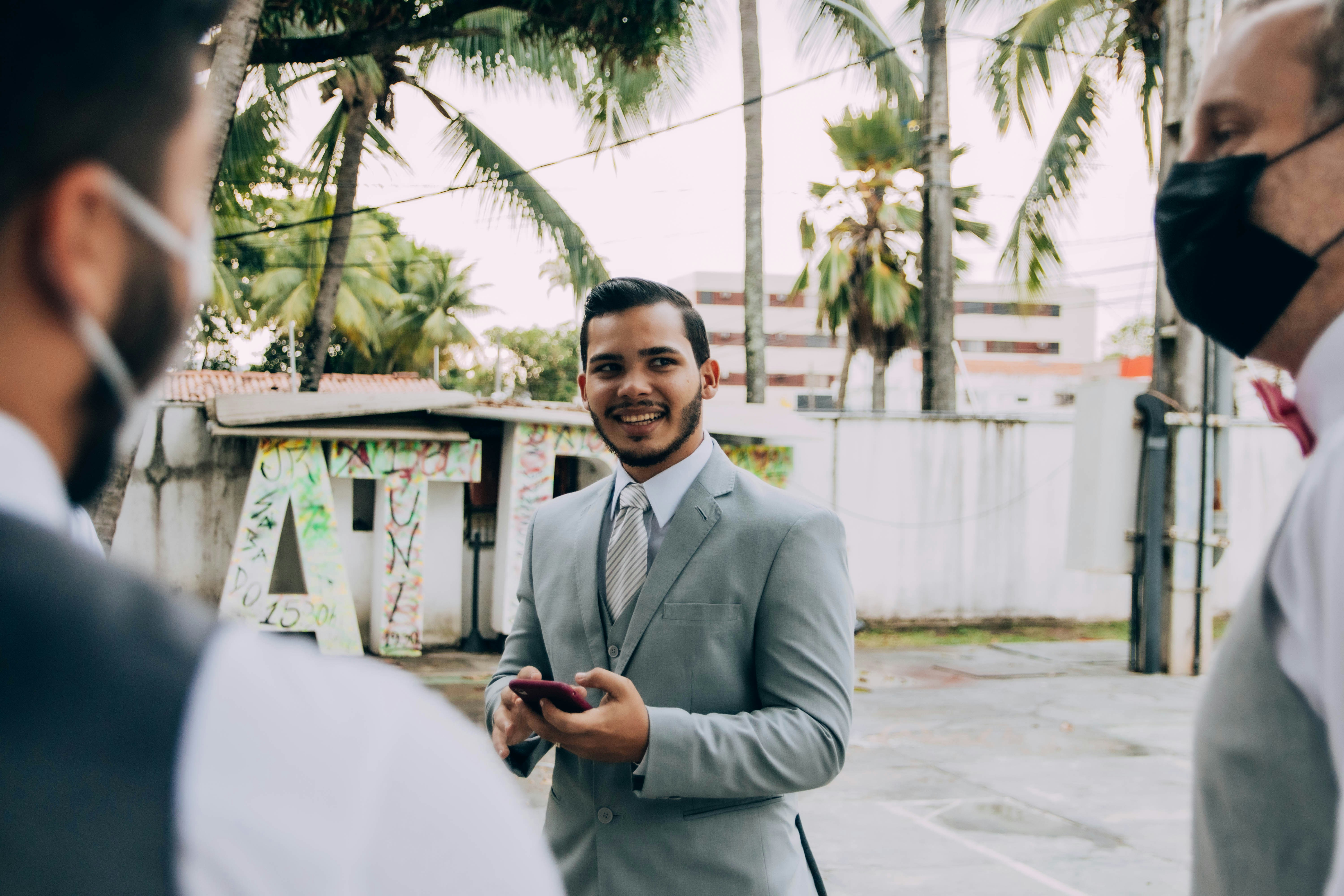 Groom in a light gray suit smiles while holding a phone, surrounded by friends in a tropical setting. The backdrop features palm trees and colorful graffiti.