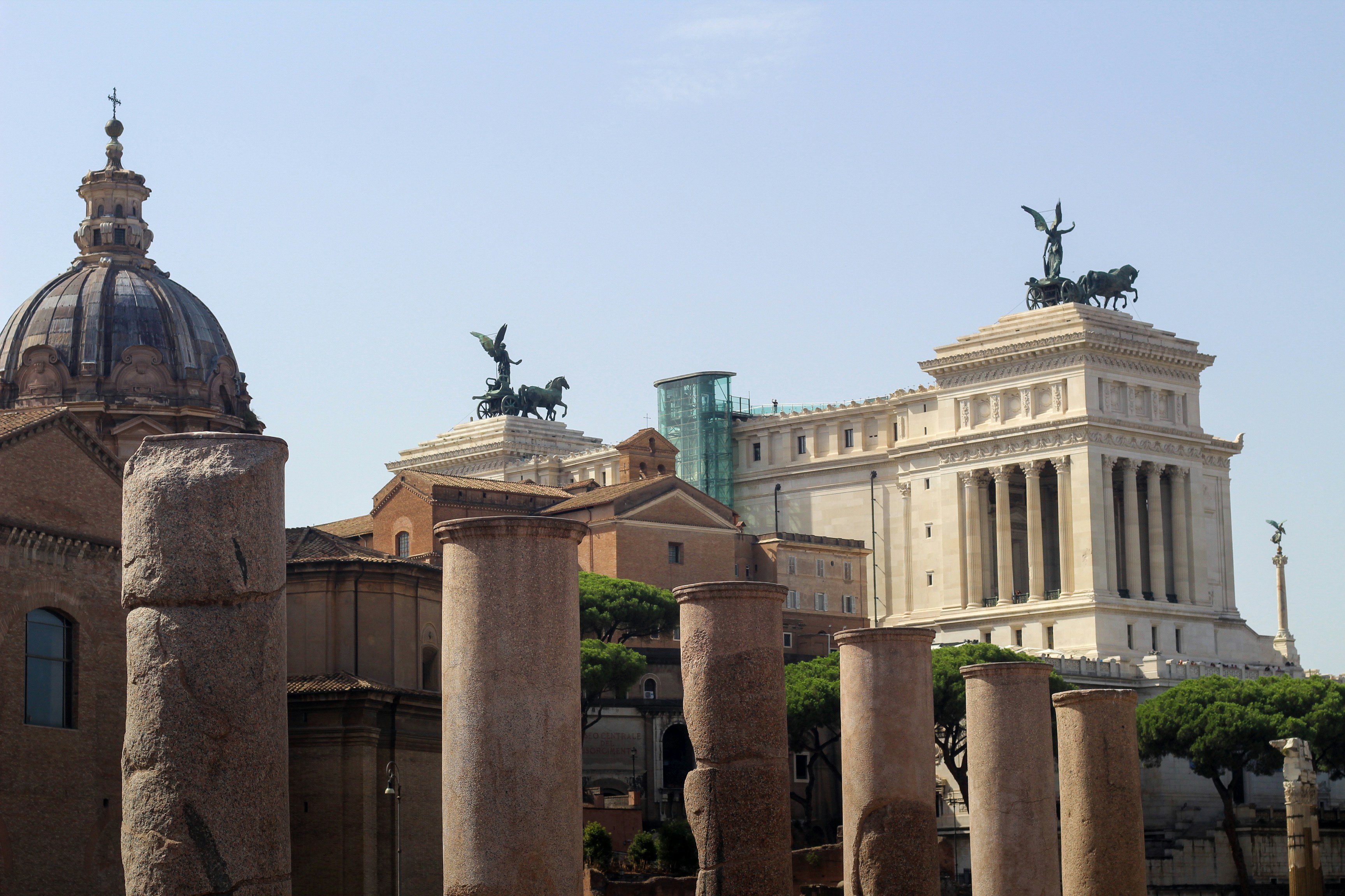 A stunning cityscape of Rome showcasing ancient Roman column ruins from the Foro Romano in the foreground, with the grand Monument to Victor Emmanuel II rising majestically in the background. This blend of historic landmarks captures the timeless beauty and layered history of Italy’s capital city. Ideal for travel, architecture, and cultural content. | Ancient roman architecture and buildings are shown.