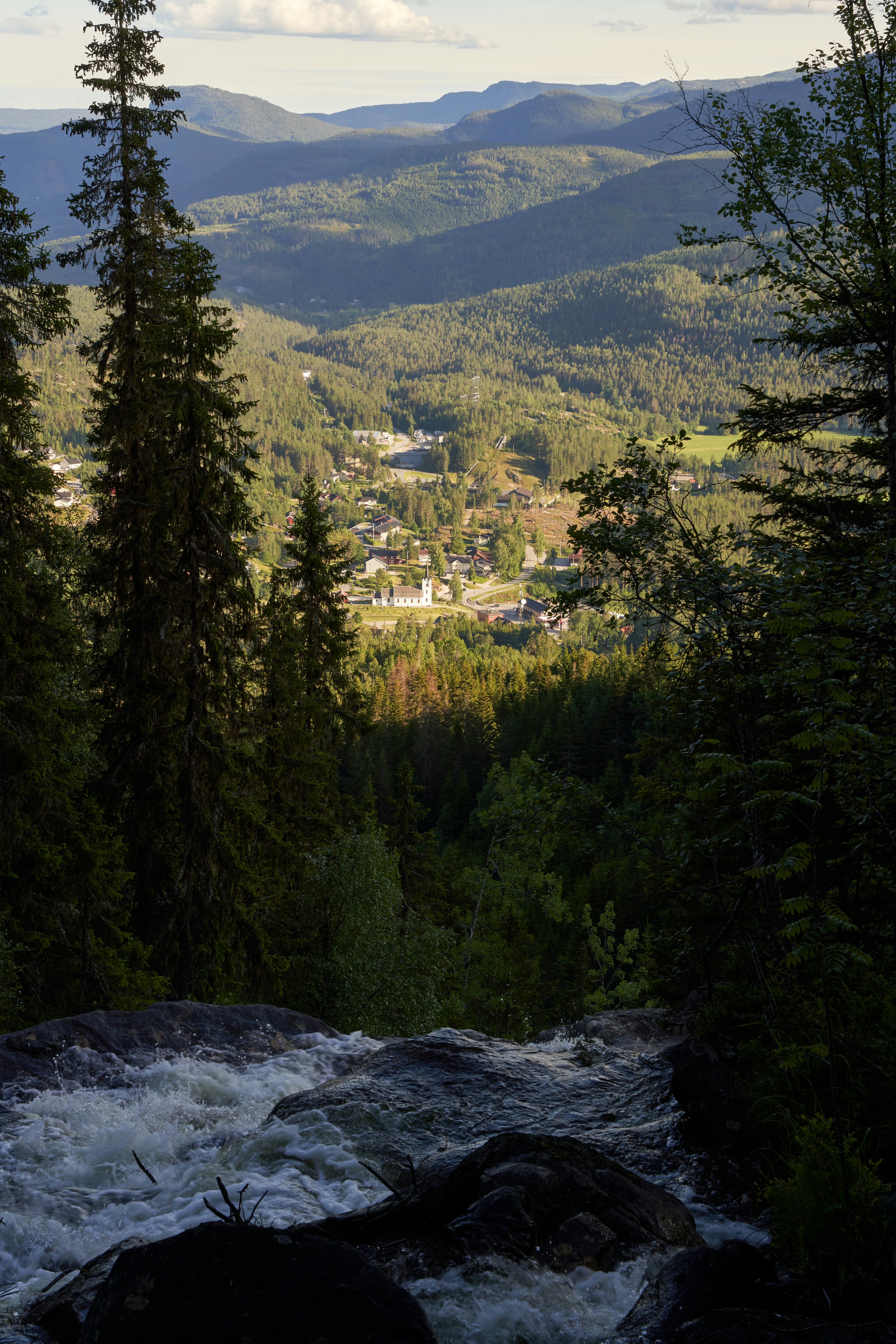 Rushing water cascades over rocks, overlooking a tranquil valley dotted with a quaint village and surrounded by lush forests and distant mountains.