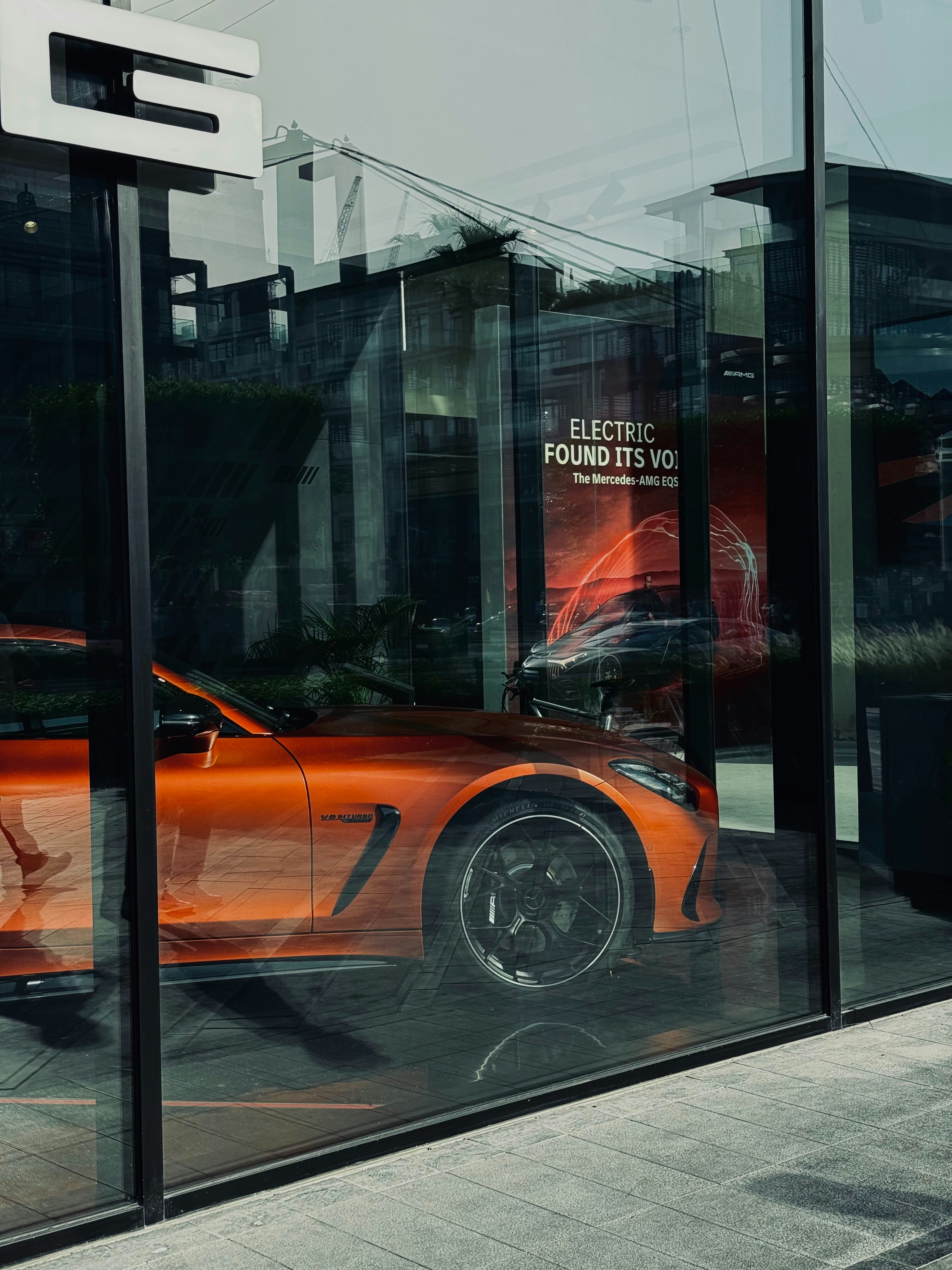 Sleek orange sports car showcased through glass, reflecting urban surroundings and a promotional display for the Mercedes-AMG EQS. 
