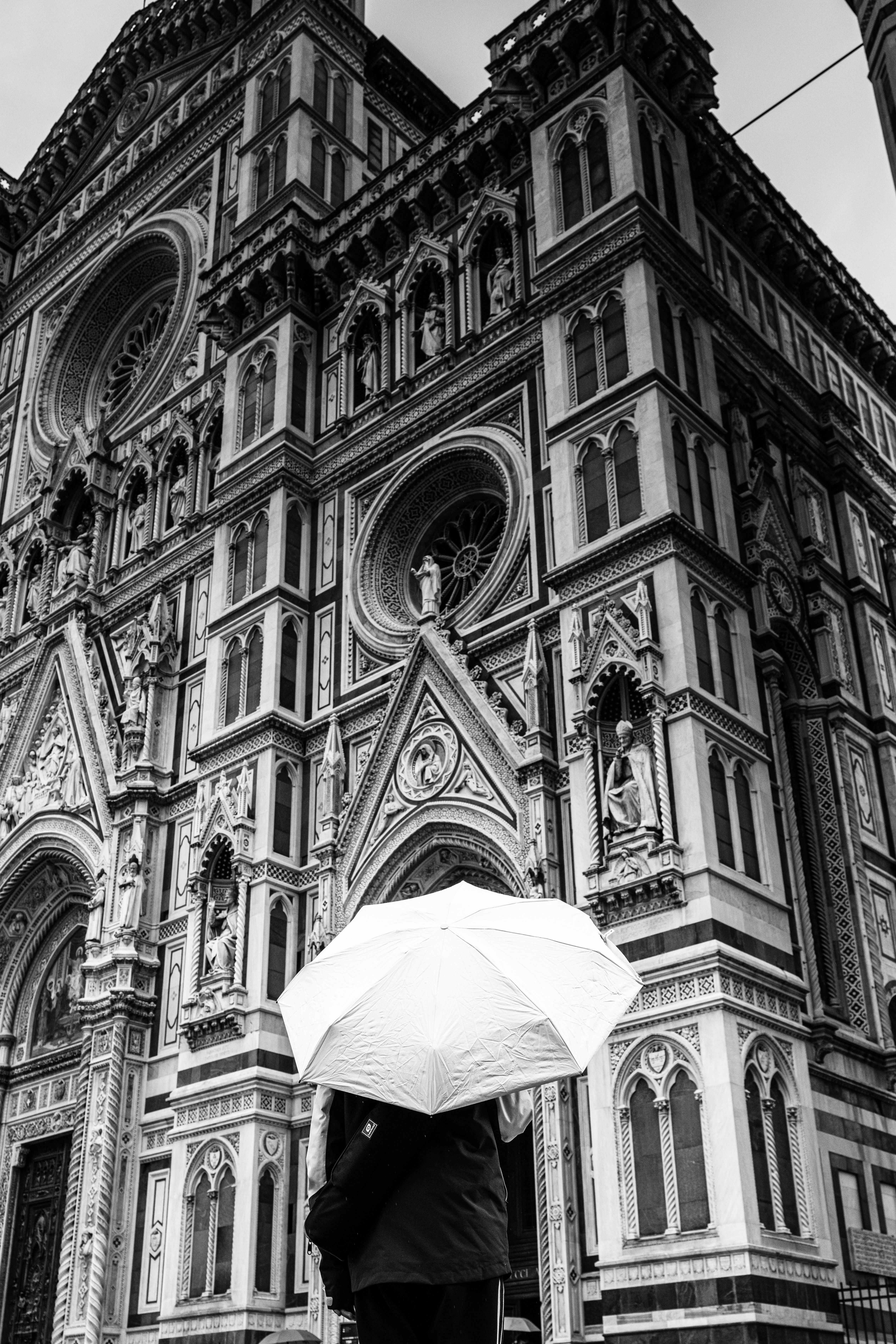A figure holding a white umbrella stands before an intricately detailed facade of a historic building. The scene is rendered in black and white, emphasizing the architectural features.
