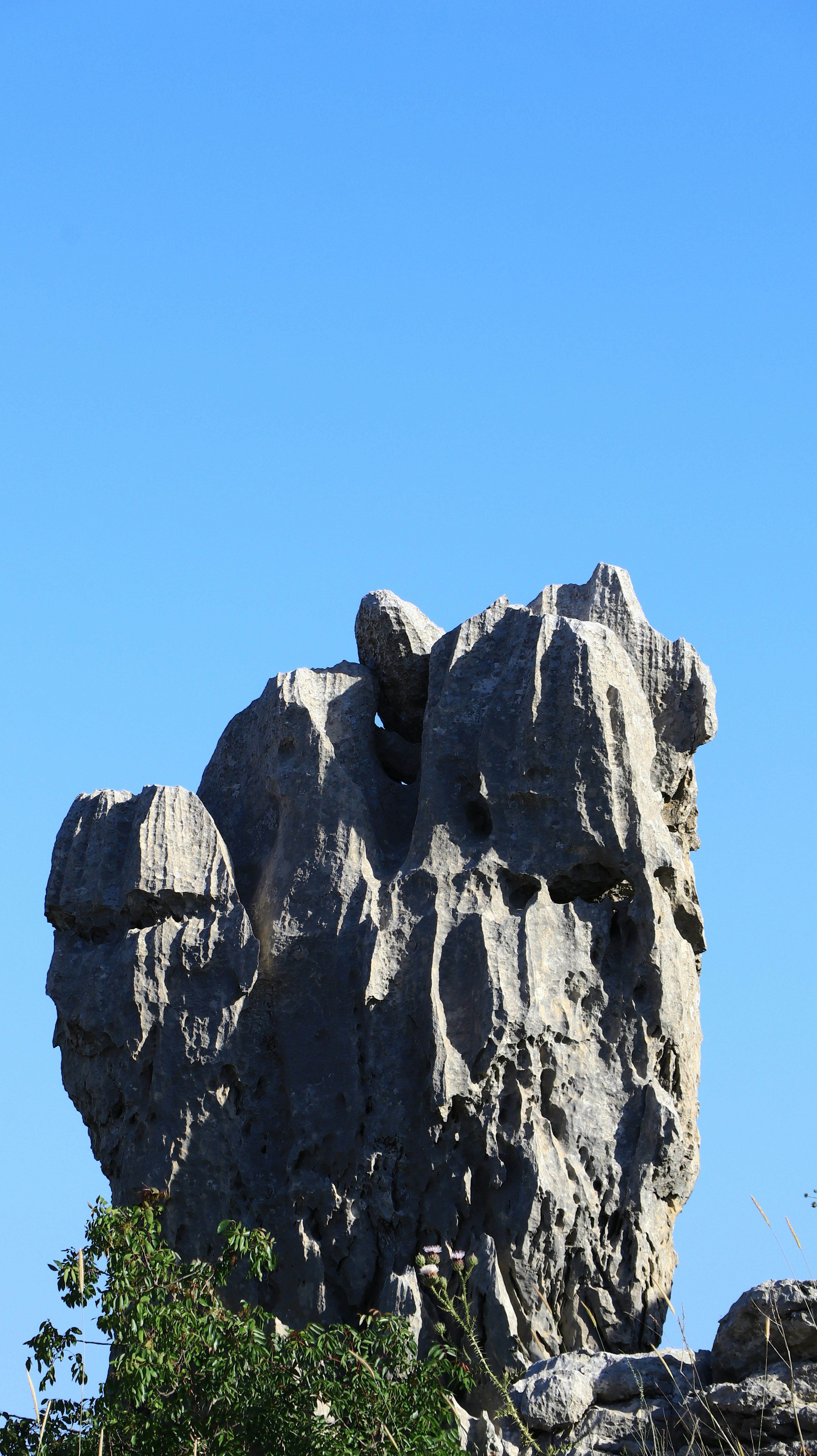A rugged rock formation resembling a sentinel stands against a clear blue sky, surrounded by sparse vegetation.