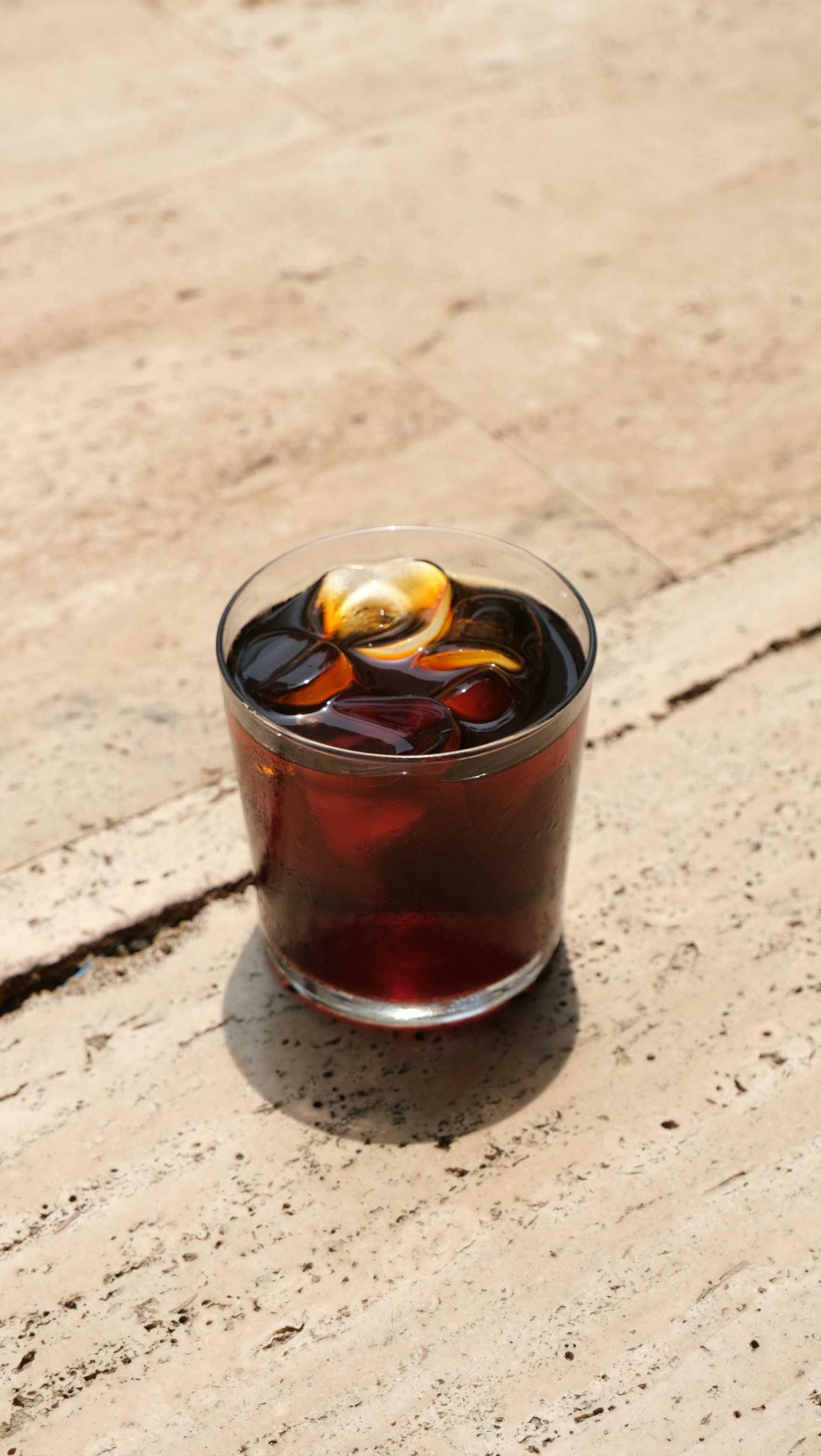 Glass of dark beverage with ice and citrus slices resting on a textured stone surface.