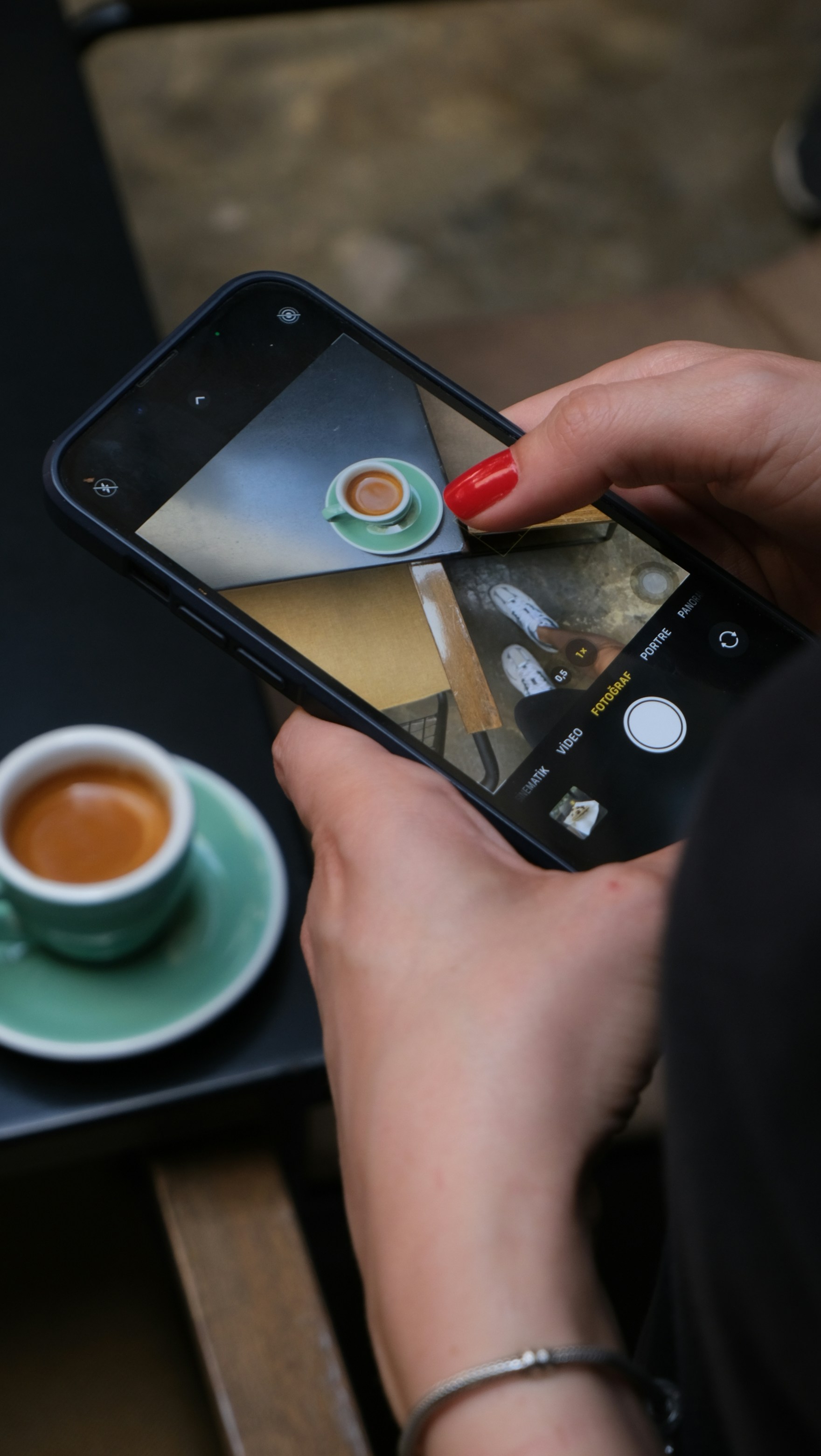 A hand holding a smartphone capturing an overhead view of an espresso cup on a wooden table, with a contrasting background. The composition emphasizes the coffee experience.