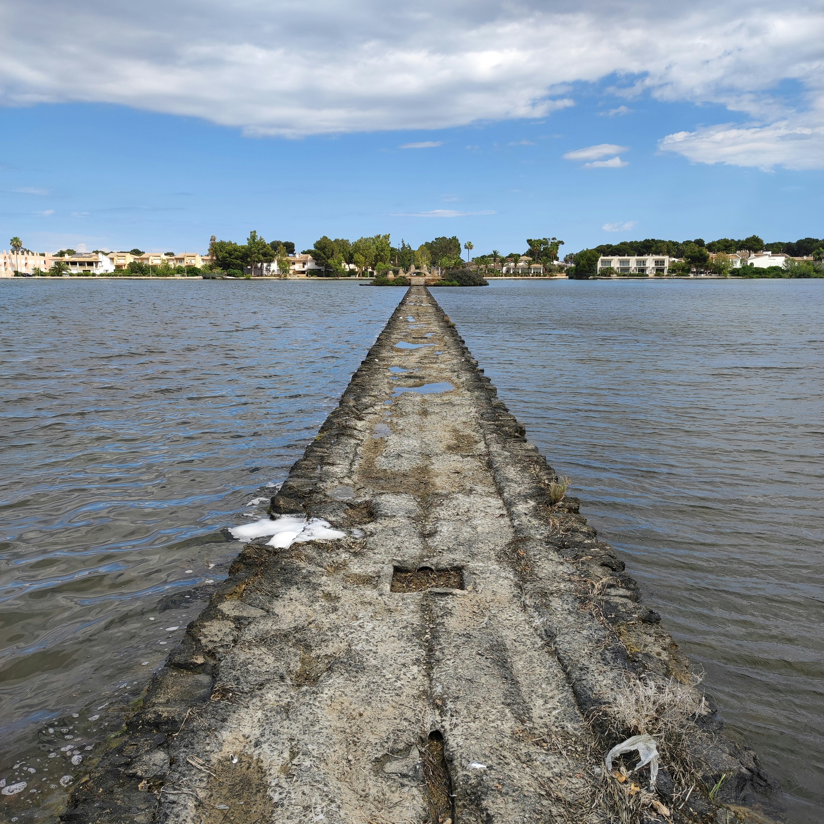 Photograph of a weathered concrete pier extending into a calm lake toward a lakeside town, with distant buildings along the shore.