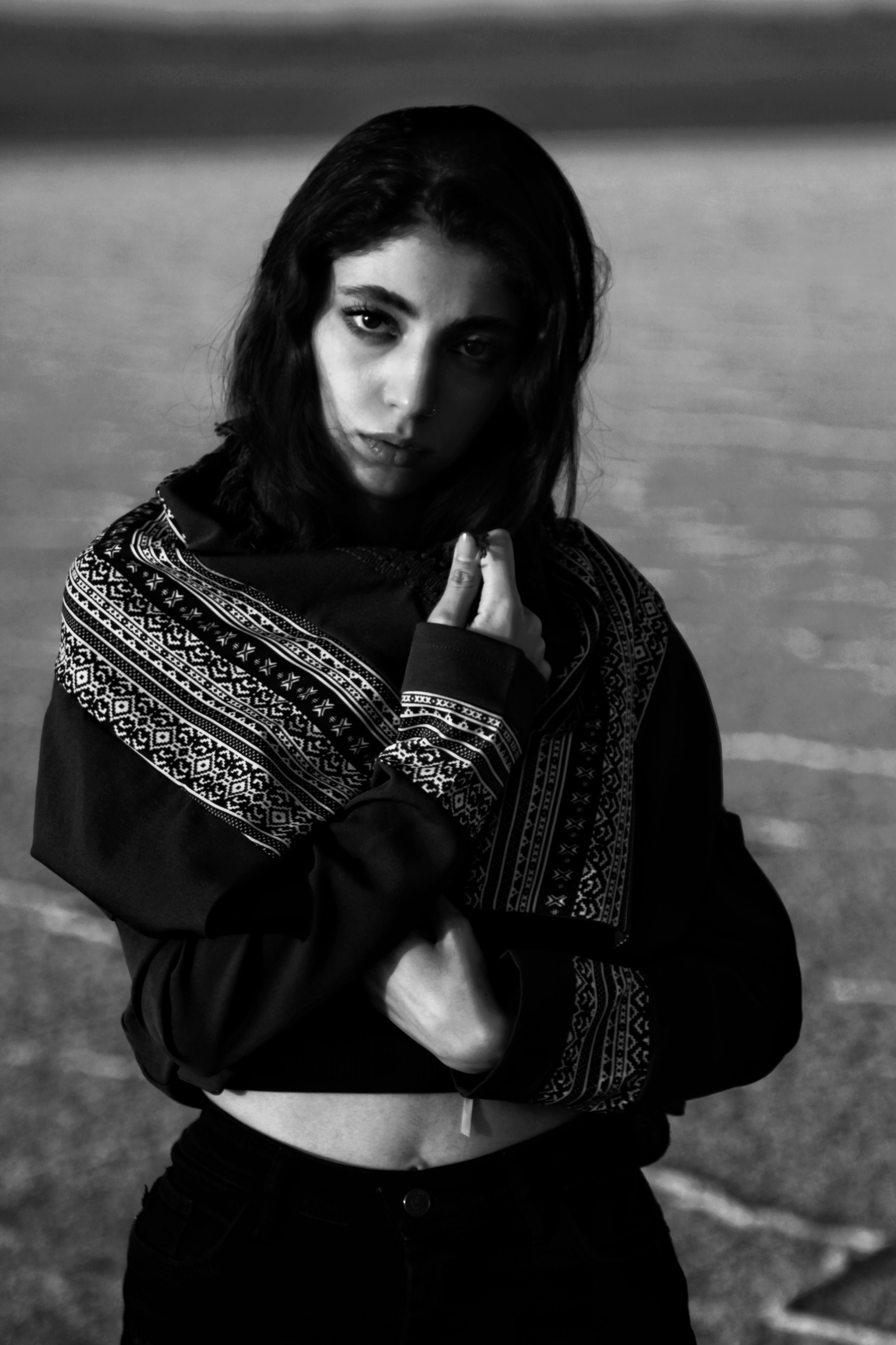 A young woman stands with a contemplative expression, draped in a patterned shawl, against a textured backdrop. The monochrome tones emphasize her thoughtful demeanor.