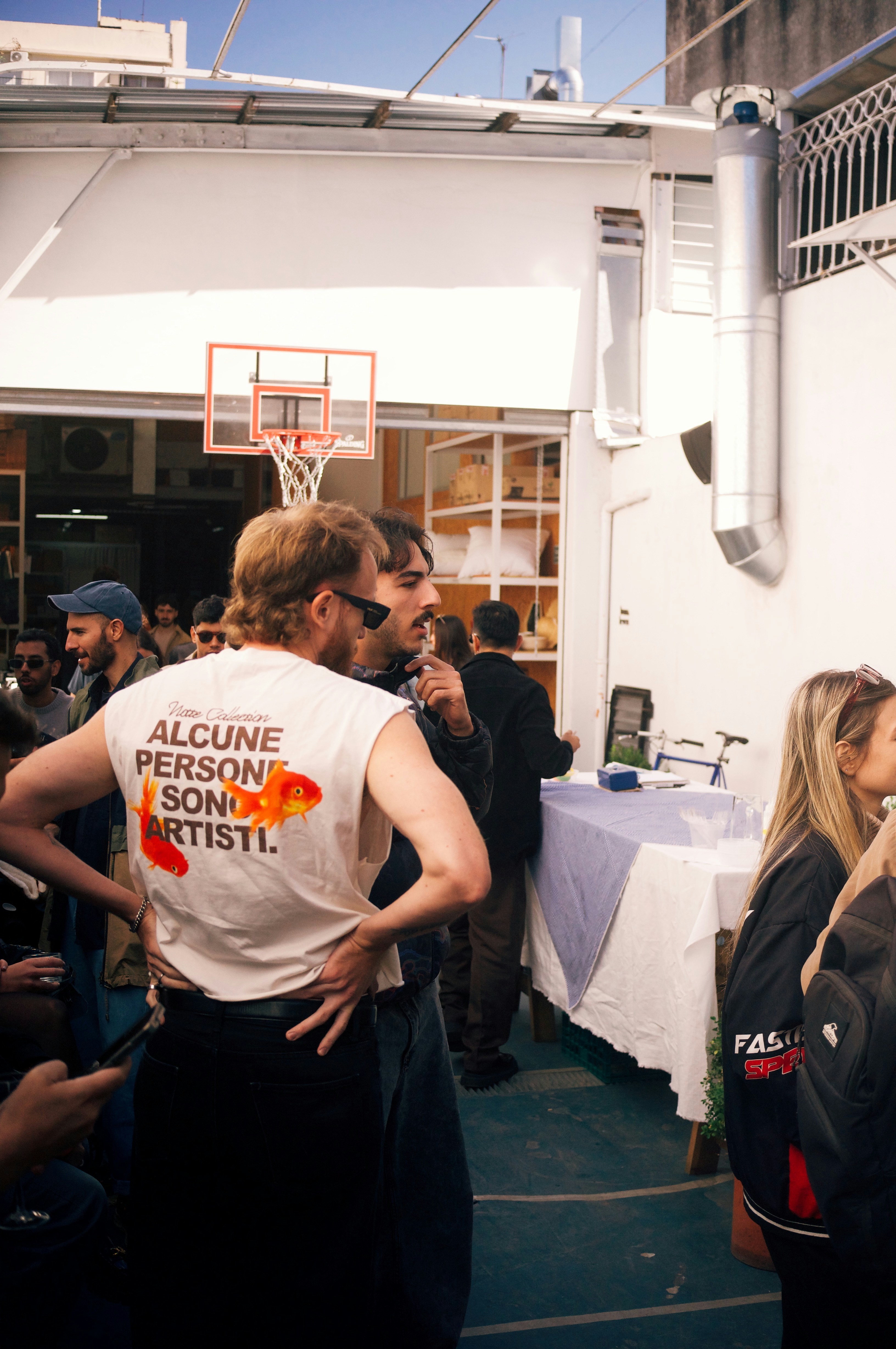 Group of people engaged in conversation in a lively setting, featuring a basketball hoop and a playful slogan on a shirt. 