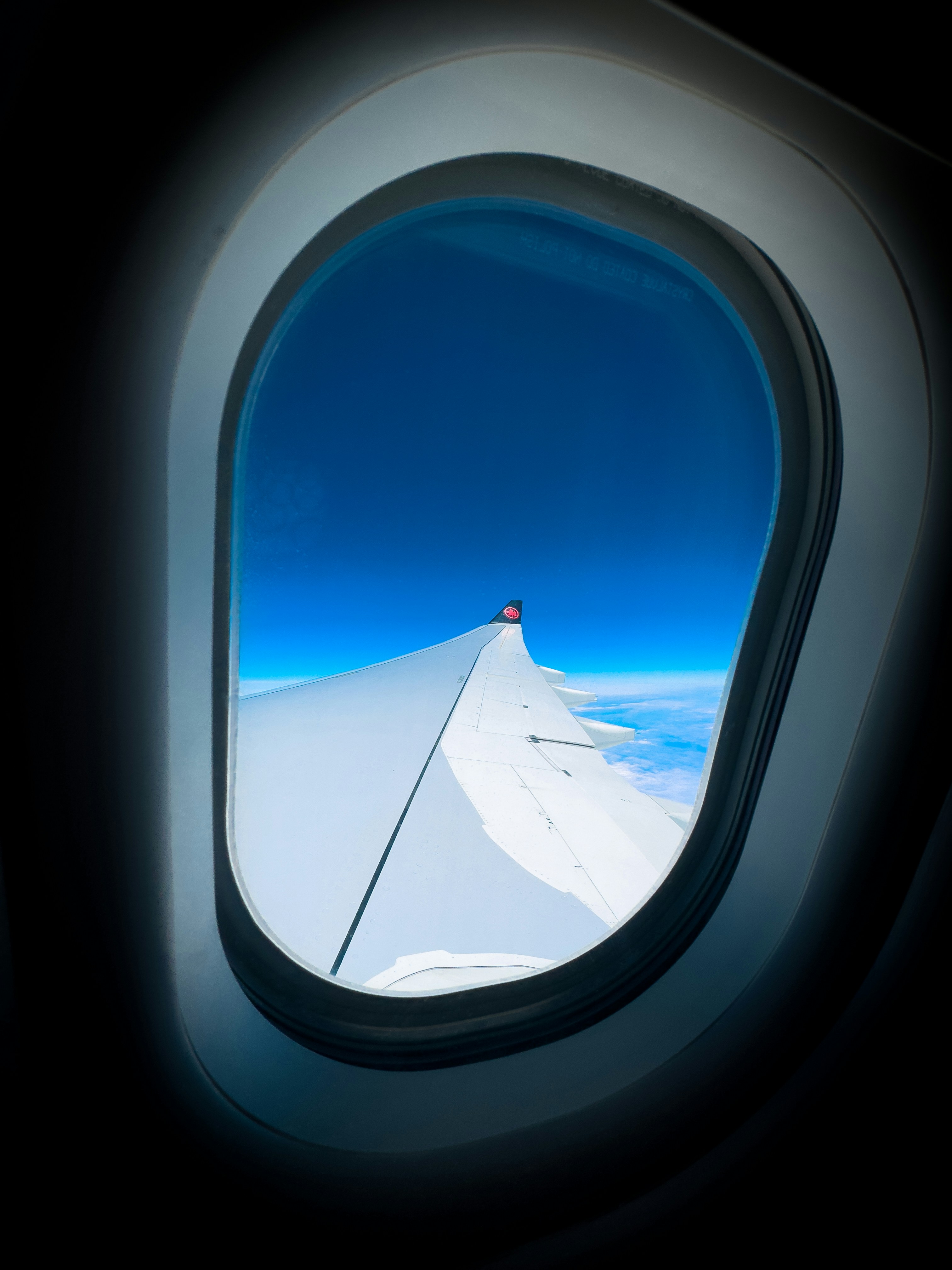 View from an airplane window showcasing an aircraft wing against a clear blue sky with wispy clouds below.