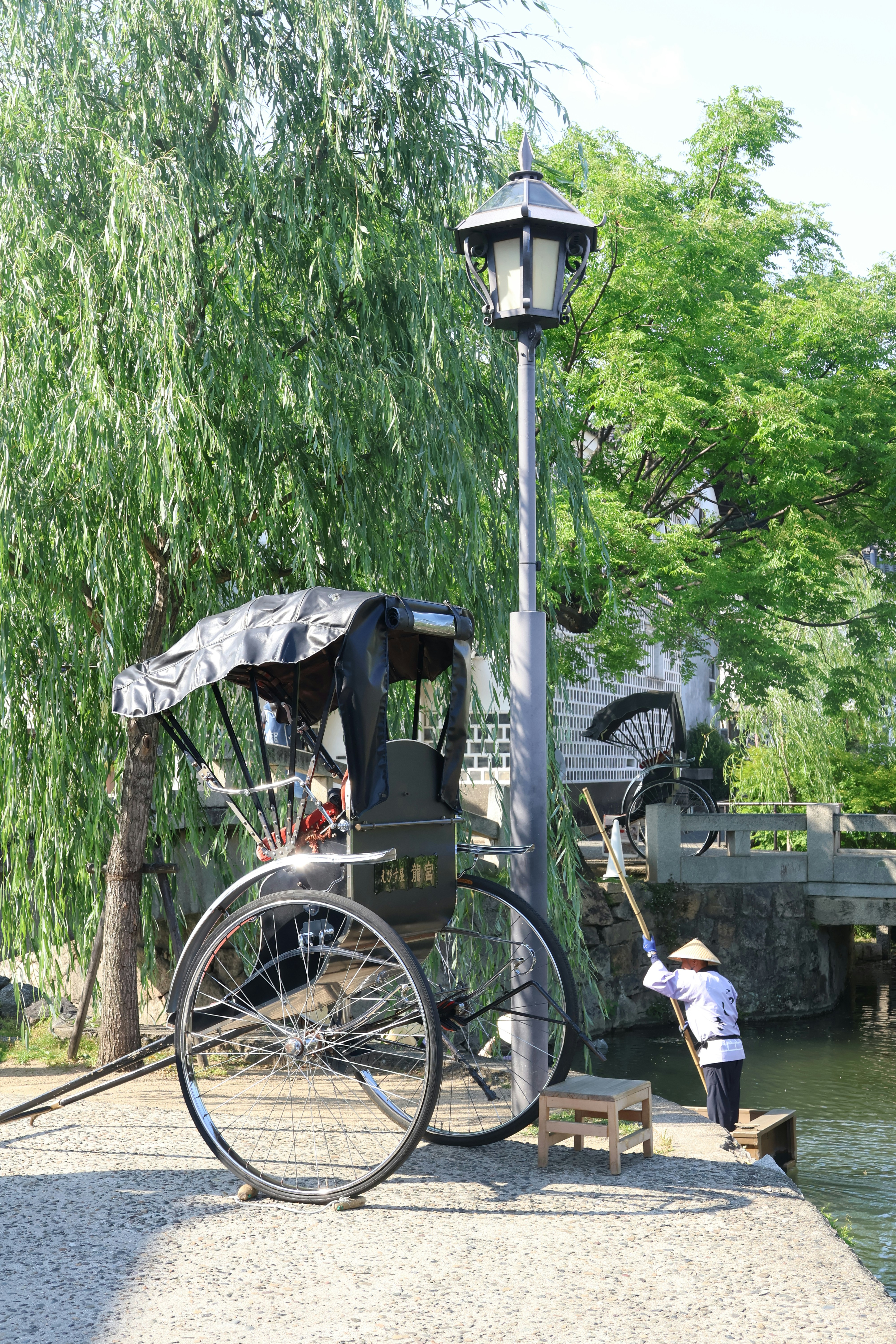 A rickshaw rests beside a serene waterway, while a figure in traditional attire tends to the surroundings. The scene captures a harmonious blend of culture and nature.