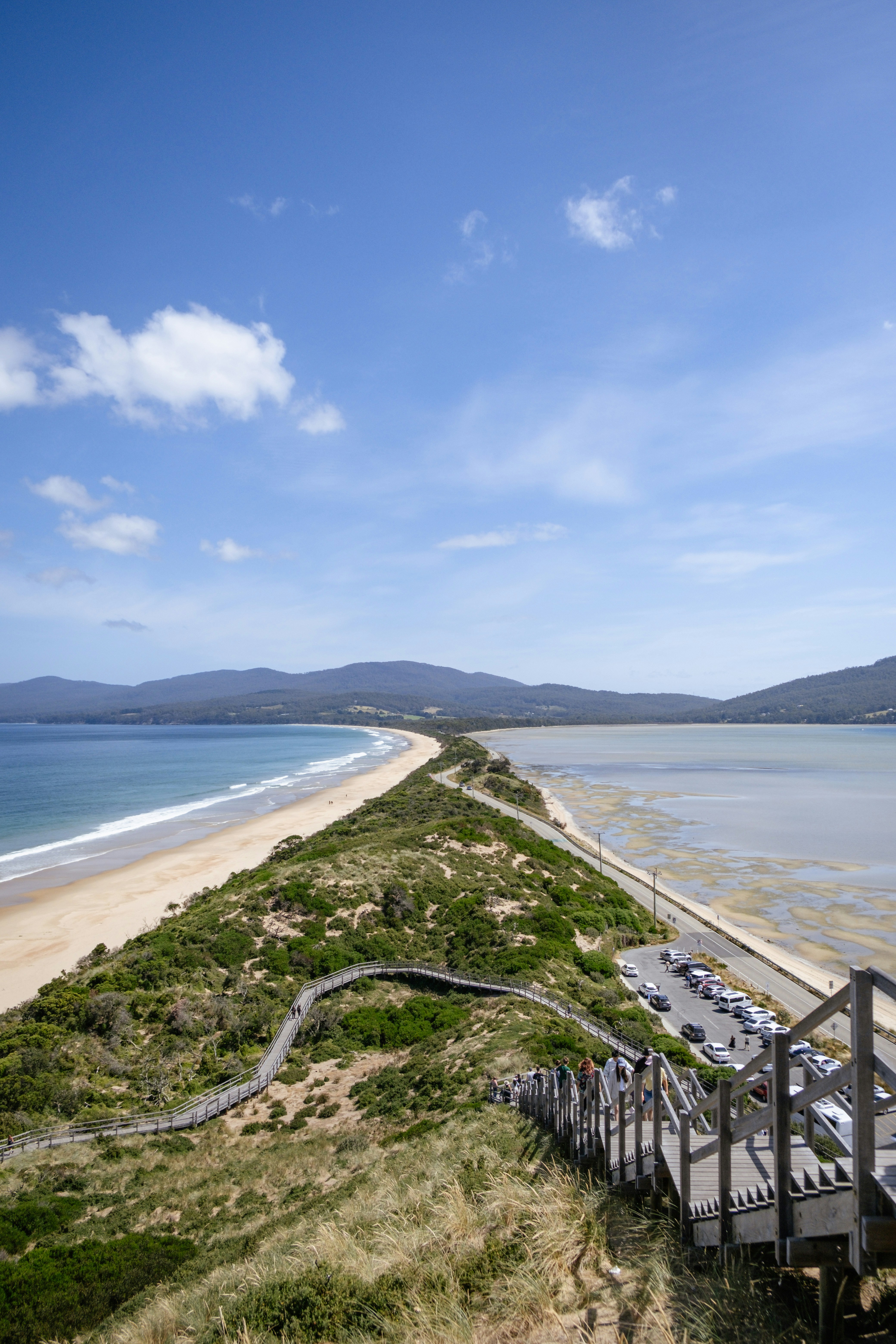 A sweeping view of a coastal landscape where a sandy beach meets lush greenery and winding pathways. The serene waters reflect the sky's hues.