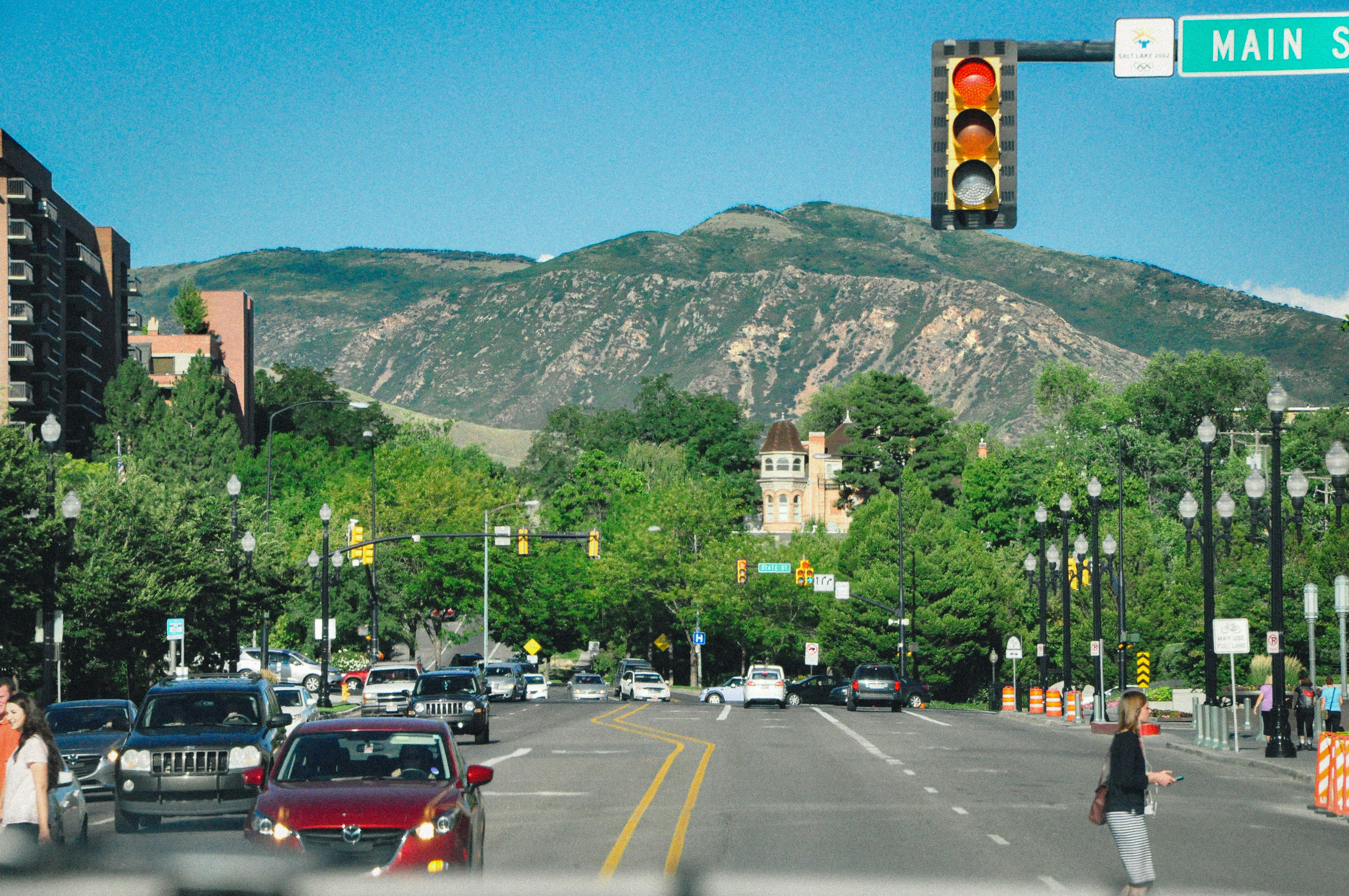 Busy city intersection with vehicles and pedestrians, framed by lush greenery and towering mountains in the background.