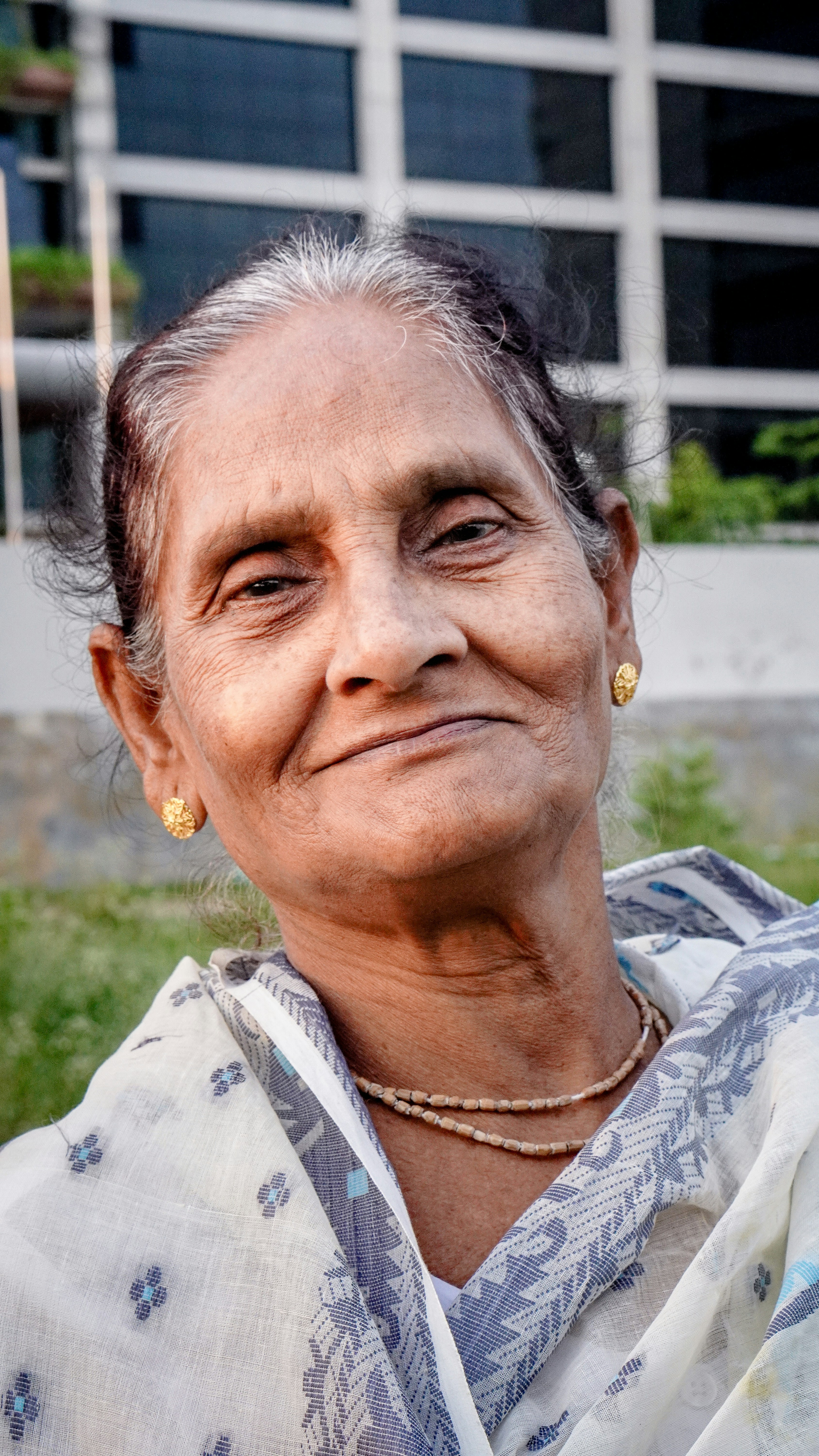 Elderly woman smiling warmly, adorned in traditional attire, set against a modern architectural backdrop.