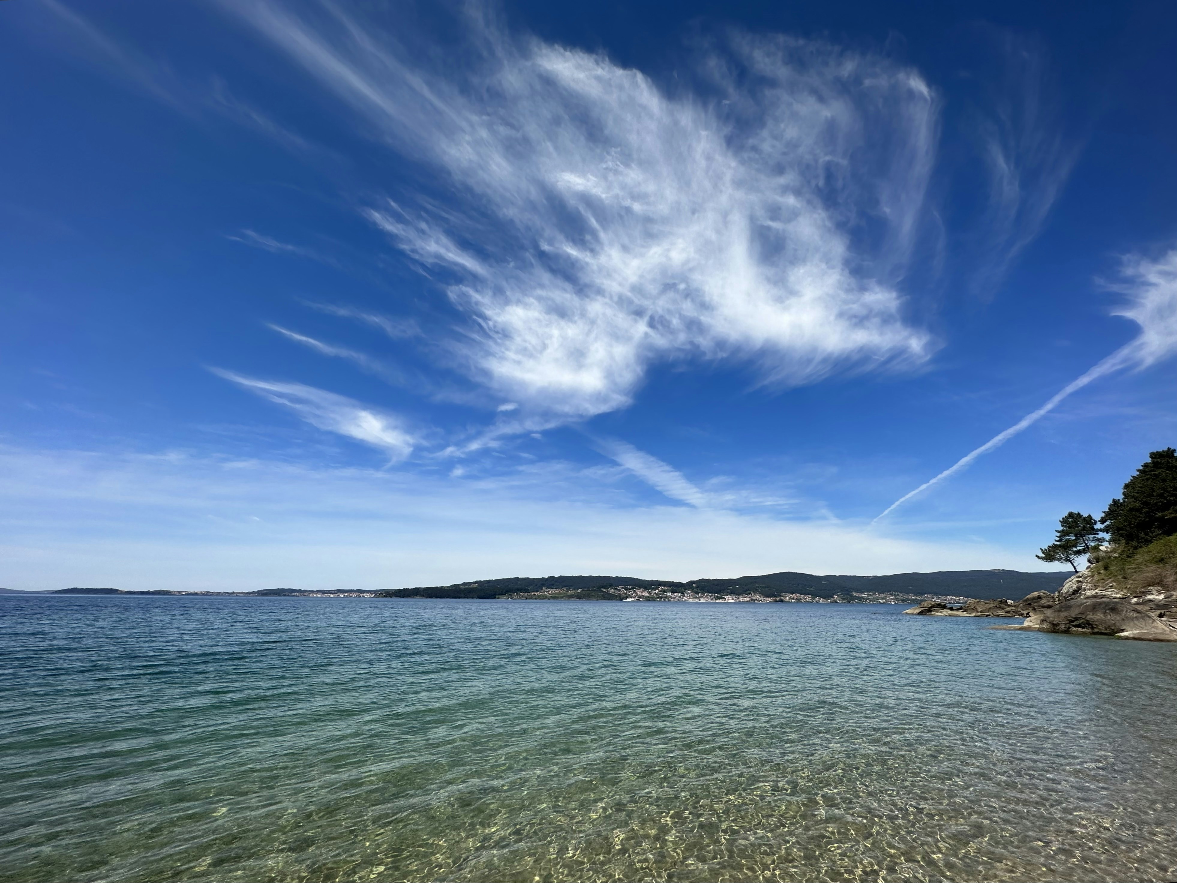 Crystal-clear waters gently lapping against a rocky shoreline under a vibrant sky with wispy clouds.