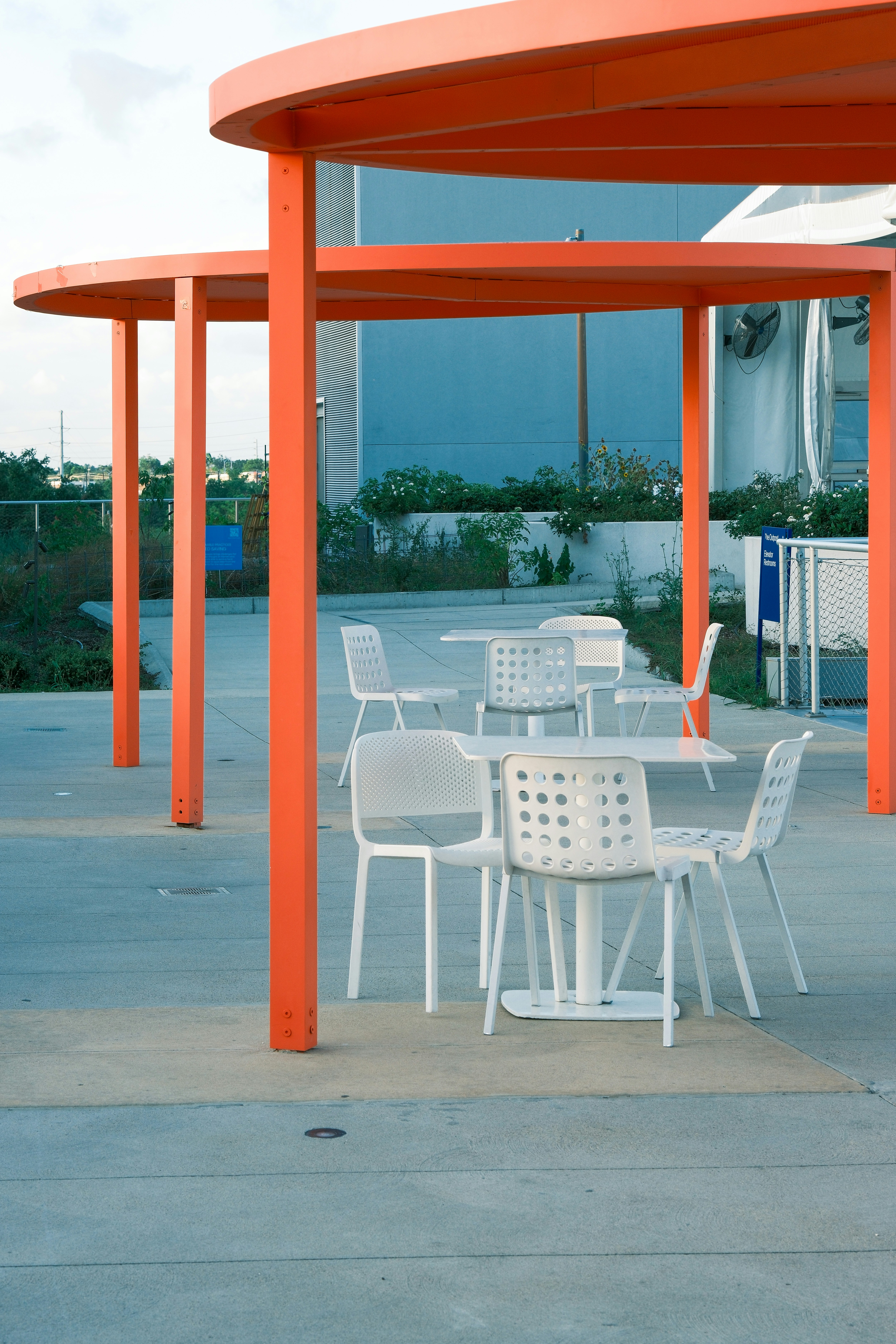 Modern white chairs and tables arranged under a bright orange pergola structure, creating a clean and geometric urban scene. | Orange structures provide shade for outdoor tables.
