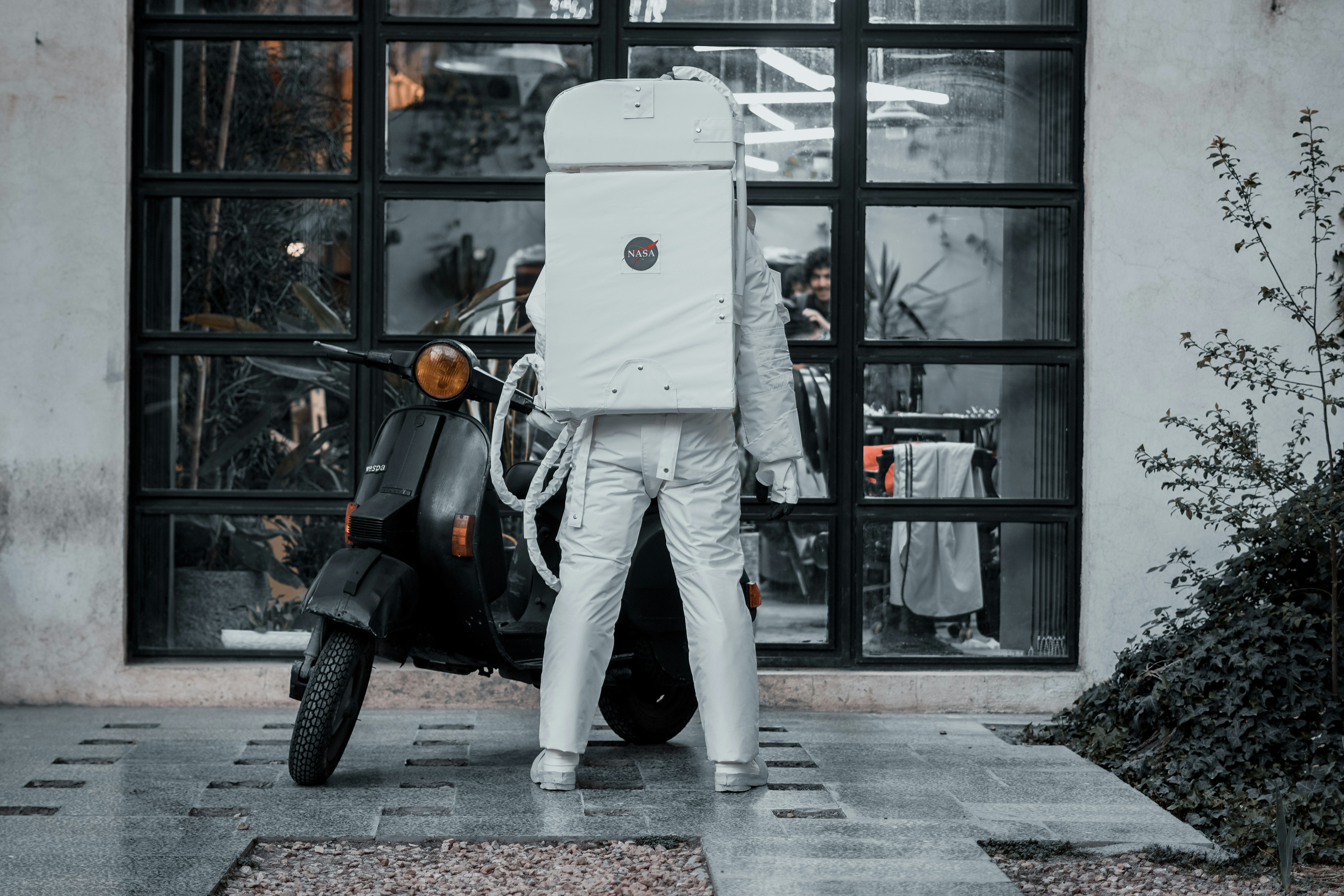 Grounded in gravity, dressed for the stars — a white-suited astronaut sits astride a black Vespa, framed by glass and steel. Retro meets sci-fi in a world where style defies orbit. NO:05 | An astronaut stands by a vintage scooter.