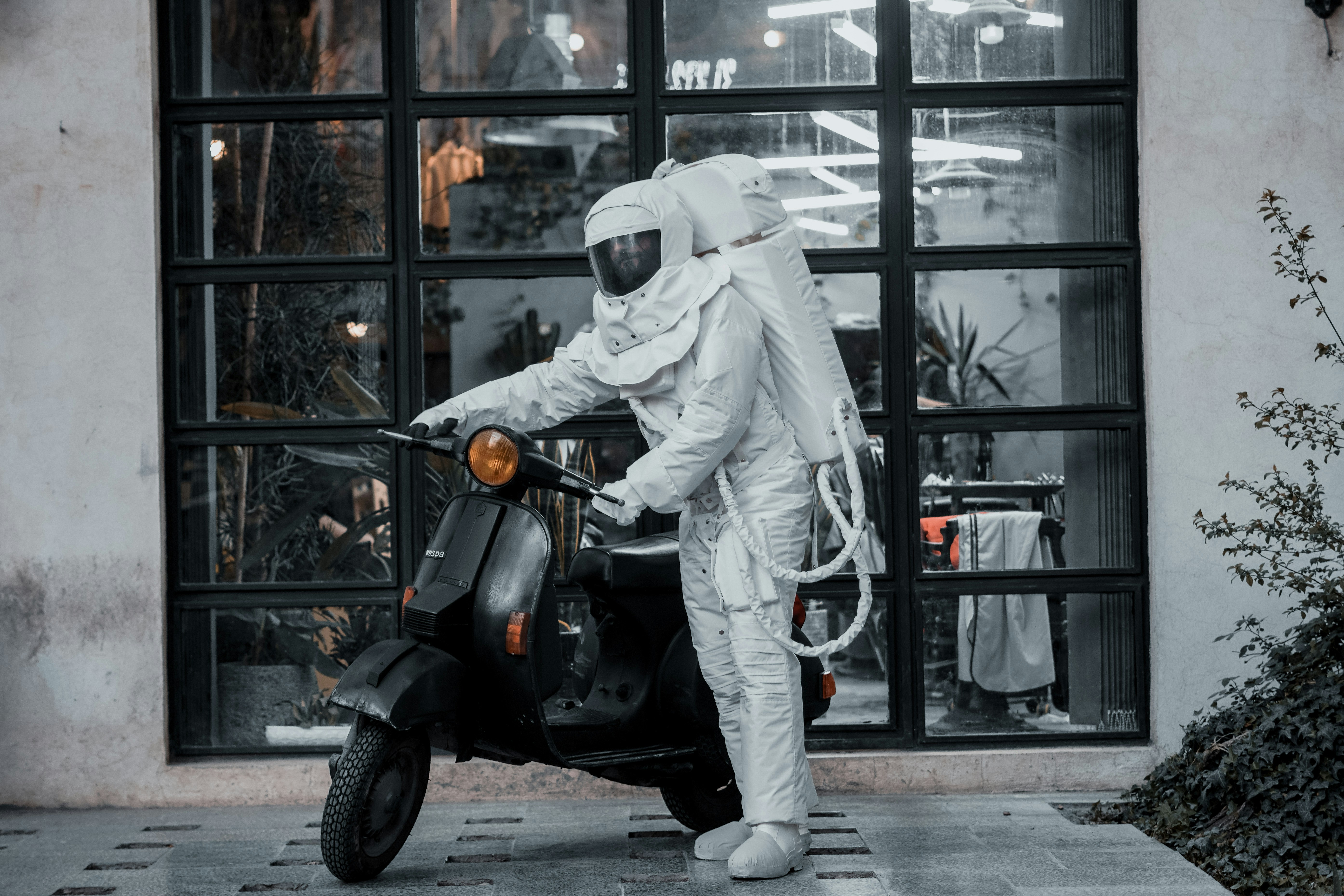 Grounded in gravity, dressed for the stars — a white-suited astronaut sits astride a black Vespa, framed by glass and steel. Retro meets sci-fi in a world where style defies orbit. NO:06 | Astronaut on a scooter ready for an adventure.
