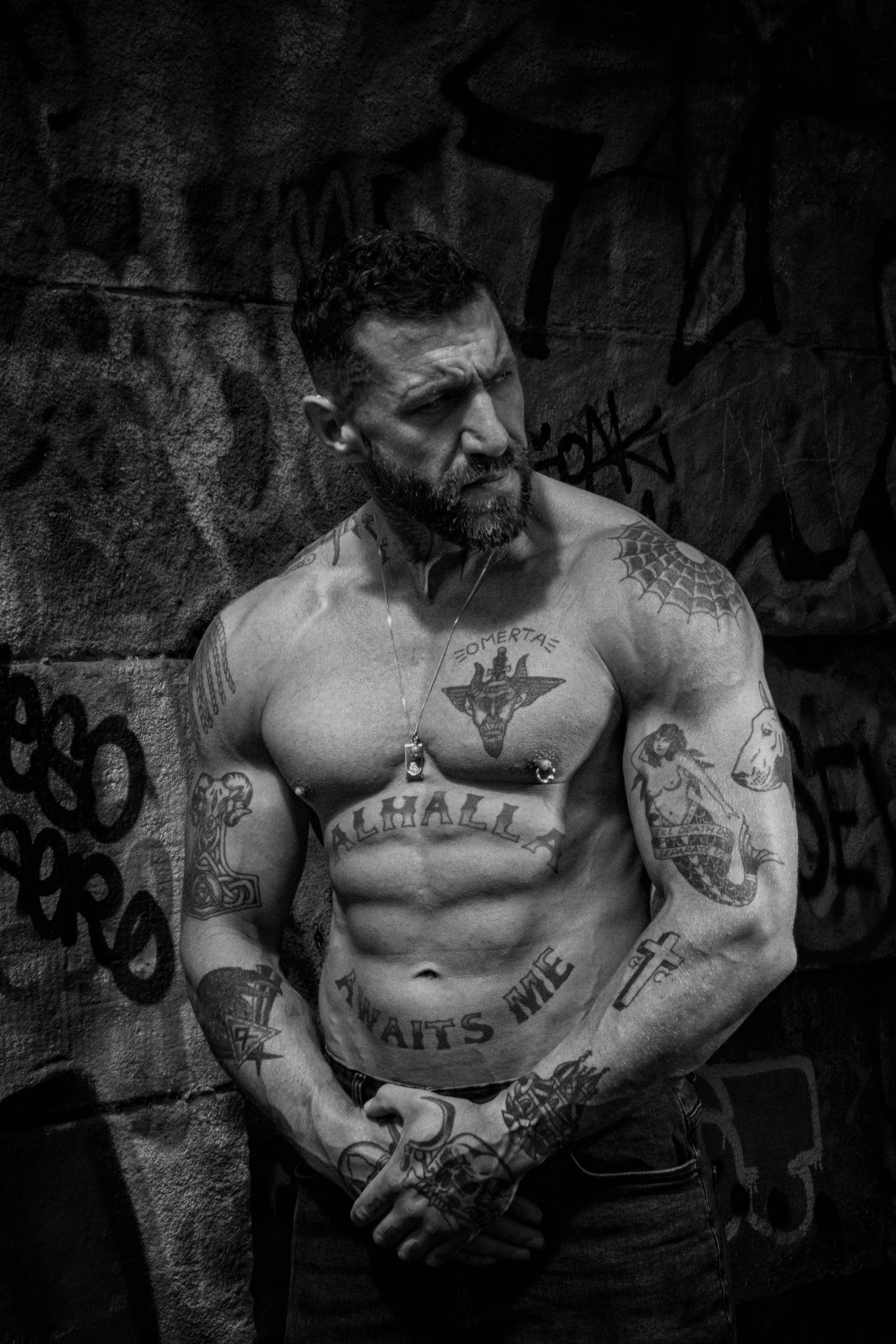 Muscular man with intricate tattoos poses against a graffiti-covered wall, exuding confidence and intensity.