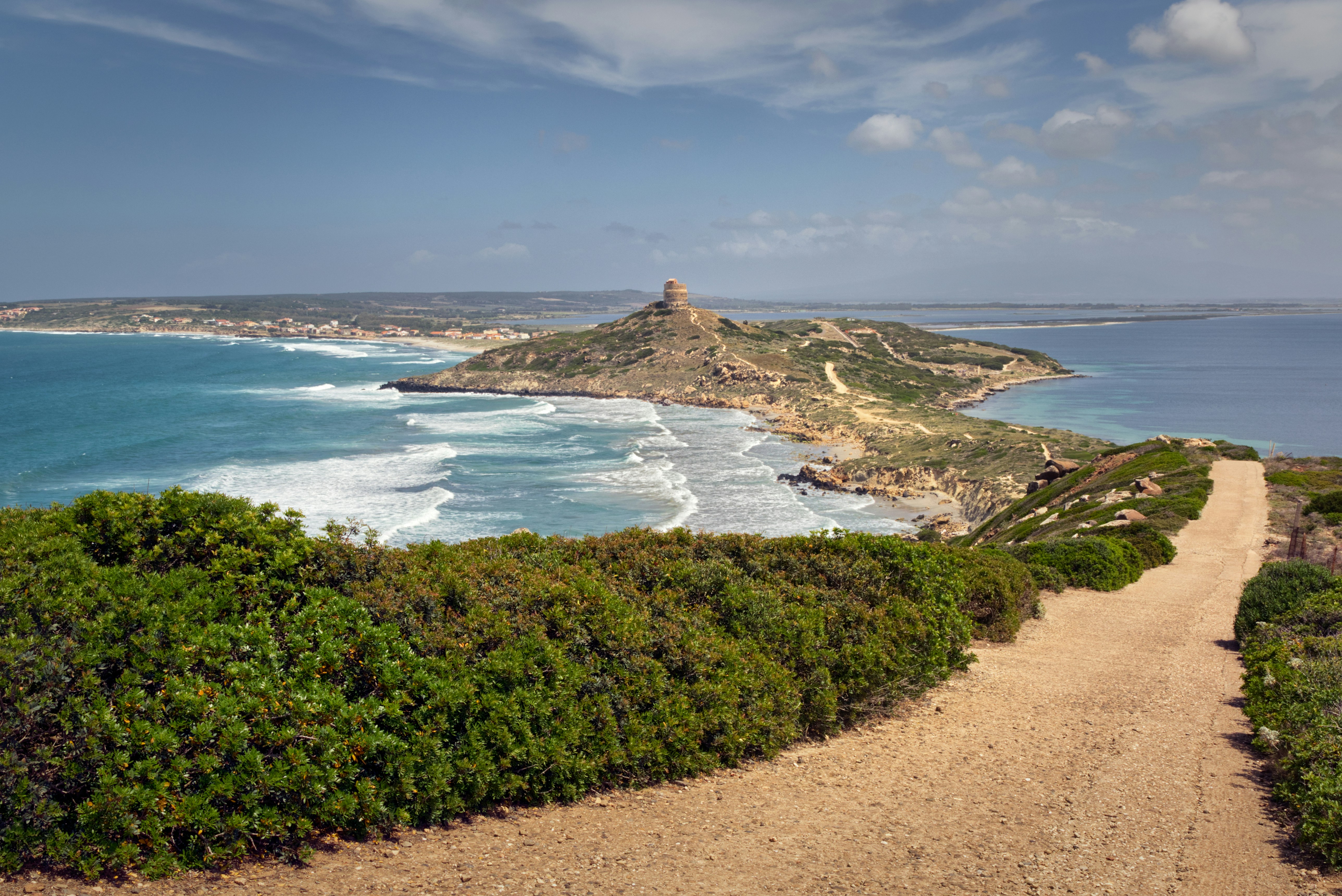 View from Capo San Marco toward Tharros | Ocean waves crash onto a sandy shore with a peninsula.