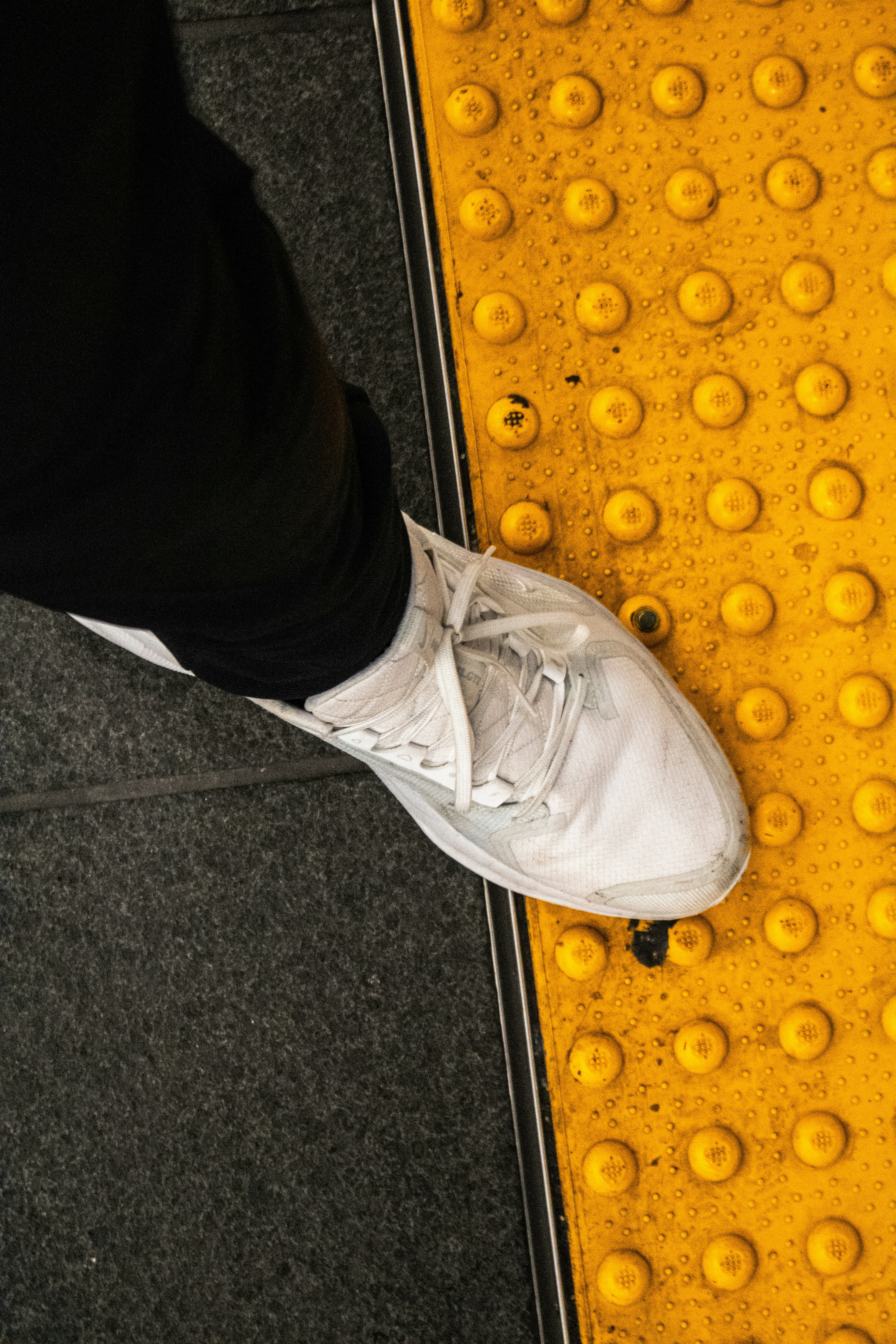 A close-up shot of a person’s foot in a white sneaker standing on the tactile yellow warning strip at a subway platform edge. Captured in an underground New York City transit station, the image highlights everyday urban textures and safety infrastructure. The bright yellow paving, designed to aid visually impaired passengers, contrasts with the dark flooring and footwear, offering a grounded look at the intersection of human movement and public design in the NYC subway system. | A person's foot steps on a tactile surface.