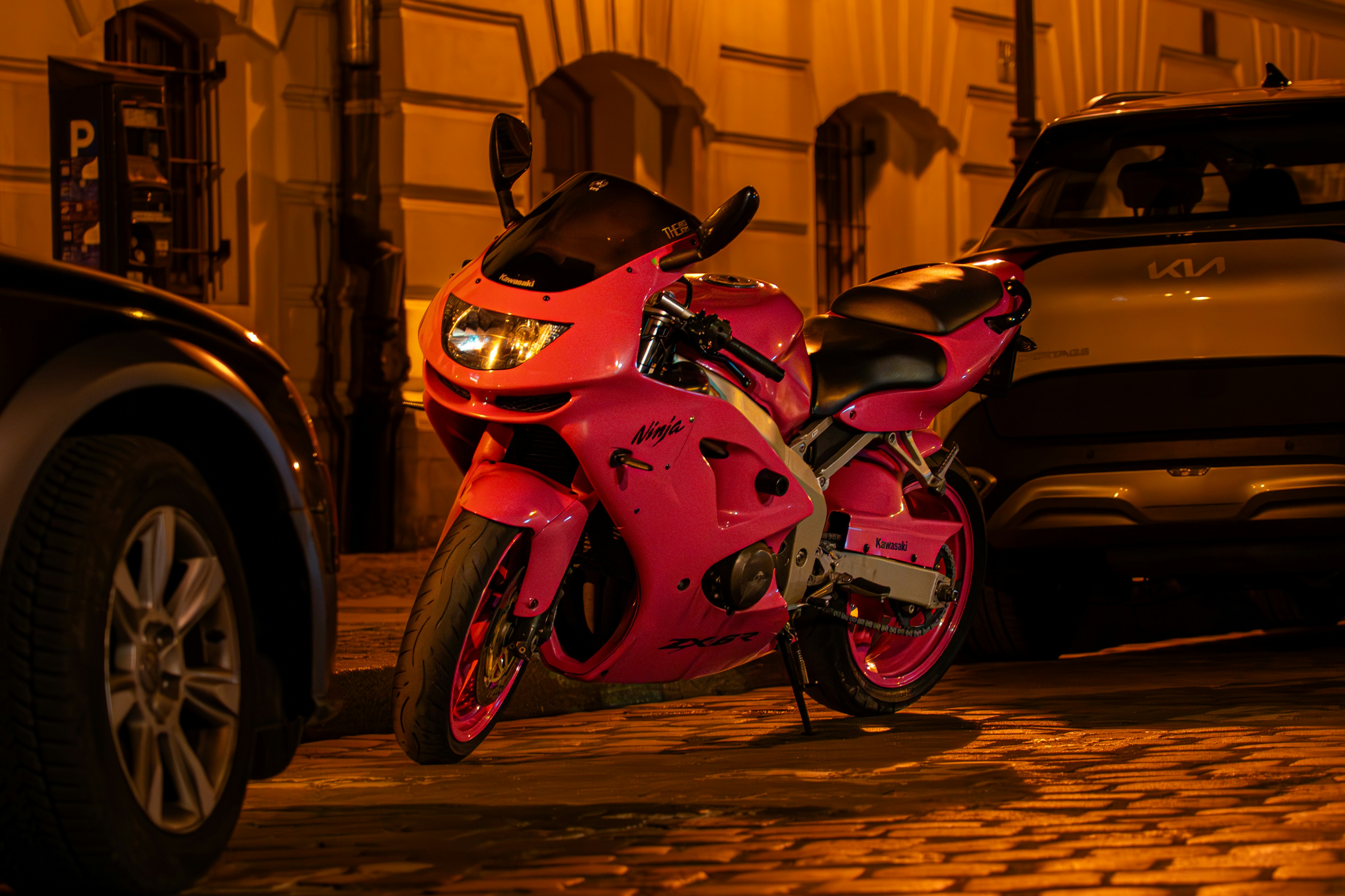 A striking pink motorcycle parked on a cobblestone street, illuminated by warm streetlights, surrounded by parked cars.