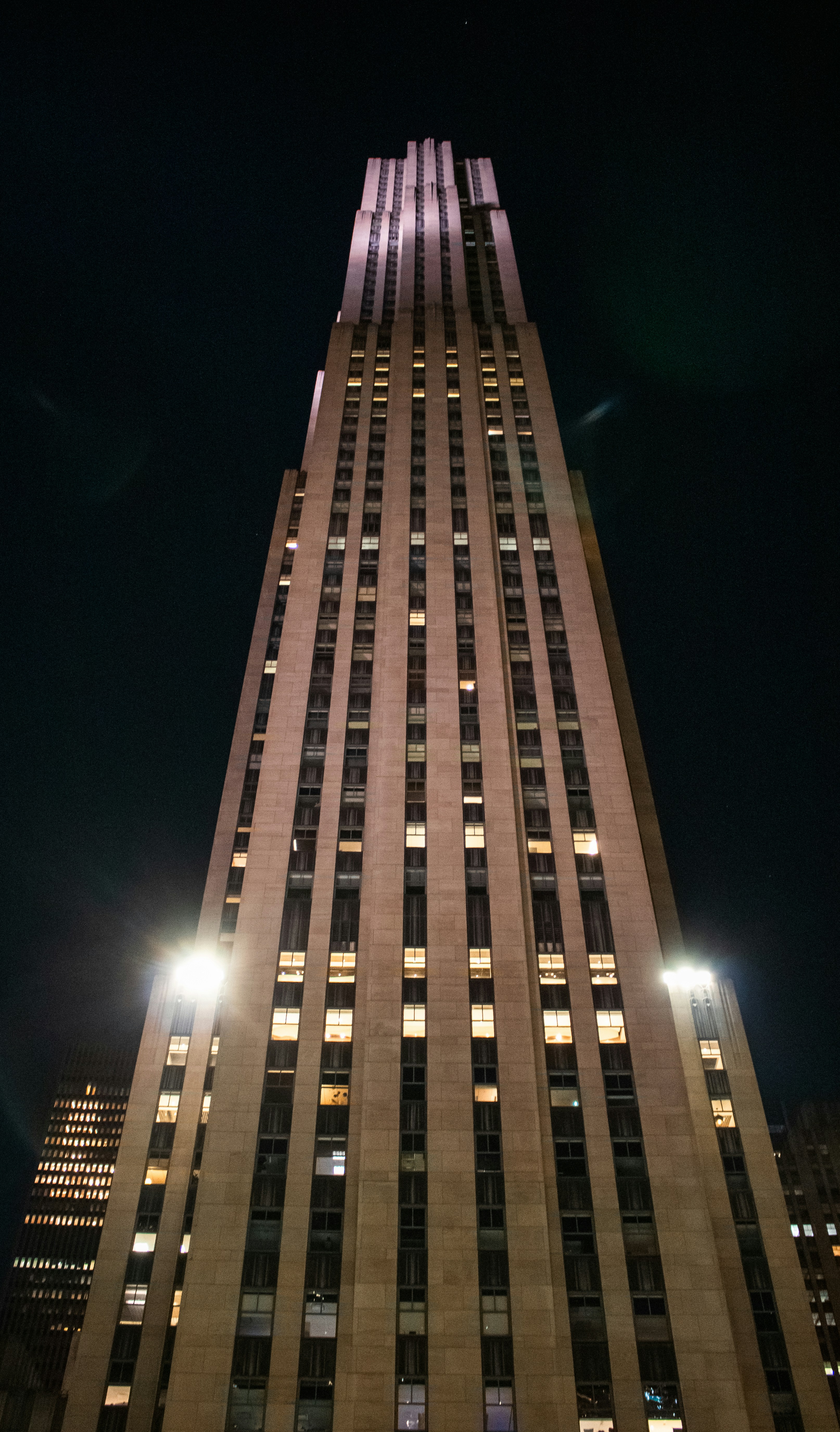 An exterior view of Rockefeller Center in Midtown Manhattan, New York City. This iconic Art Deco complex is a major landmark known for its central plaza, towering buildings, and historic significance. The site draws both tourists and locals year-round and hosts seasonal events like the famous Christmas tree lighting. | A tall building illuminated at night.