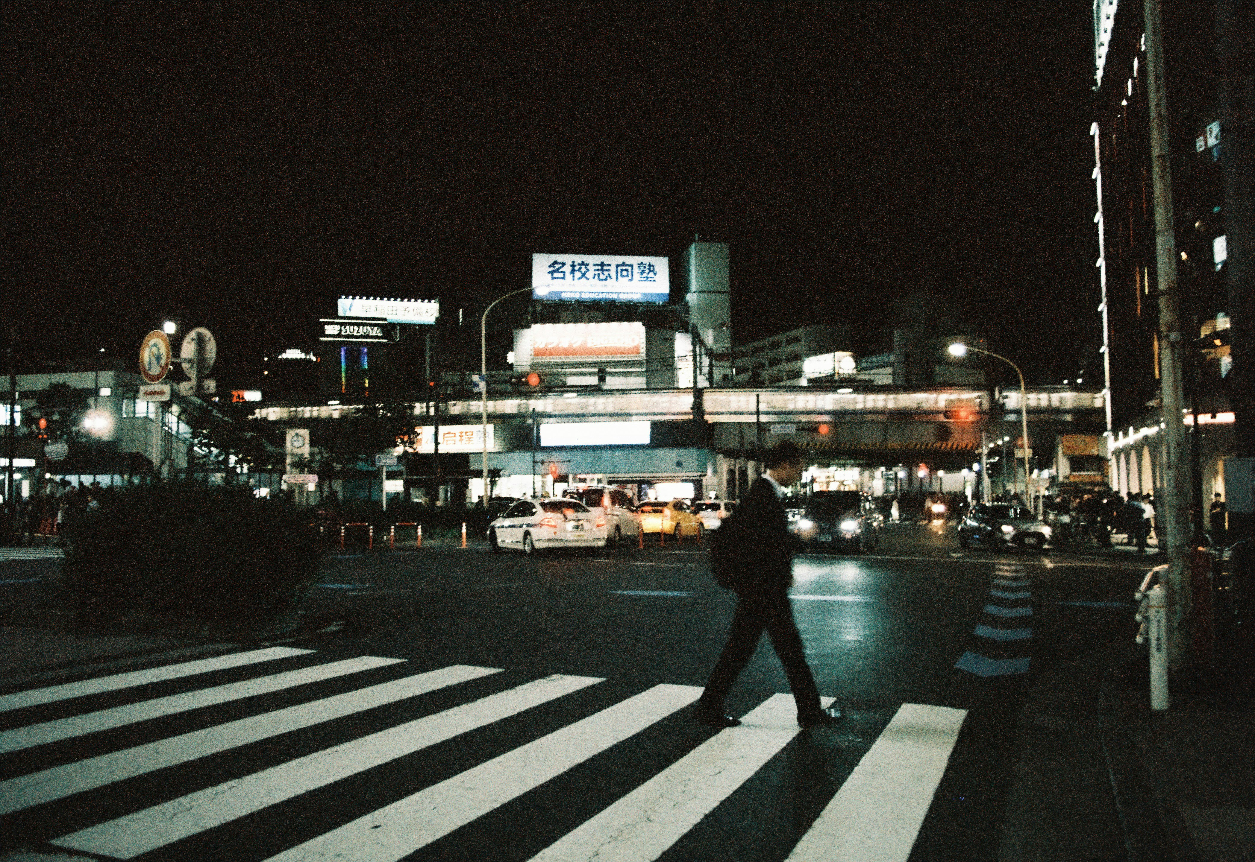 A lone figure crosses a striped crosswalk under the glow of neon signs in a bustling city at night.