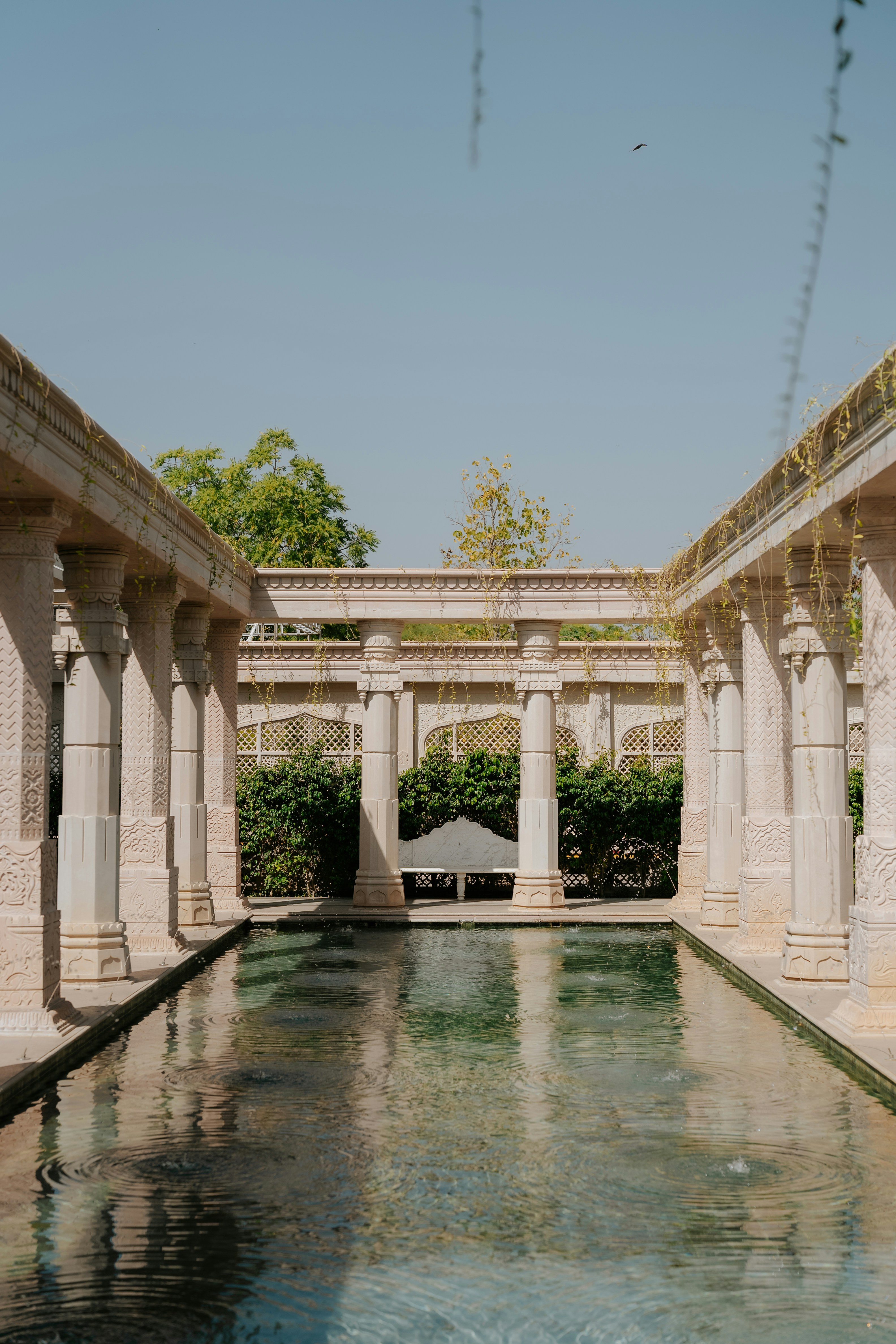 Intricate stone columns frame a tranquil waterway, reflecting the lush greenery and blue sky above.