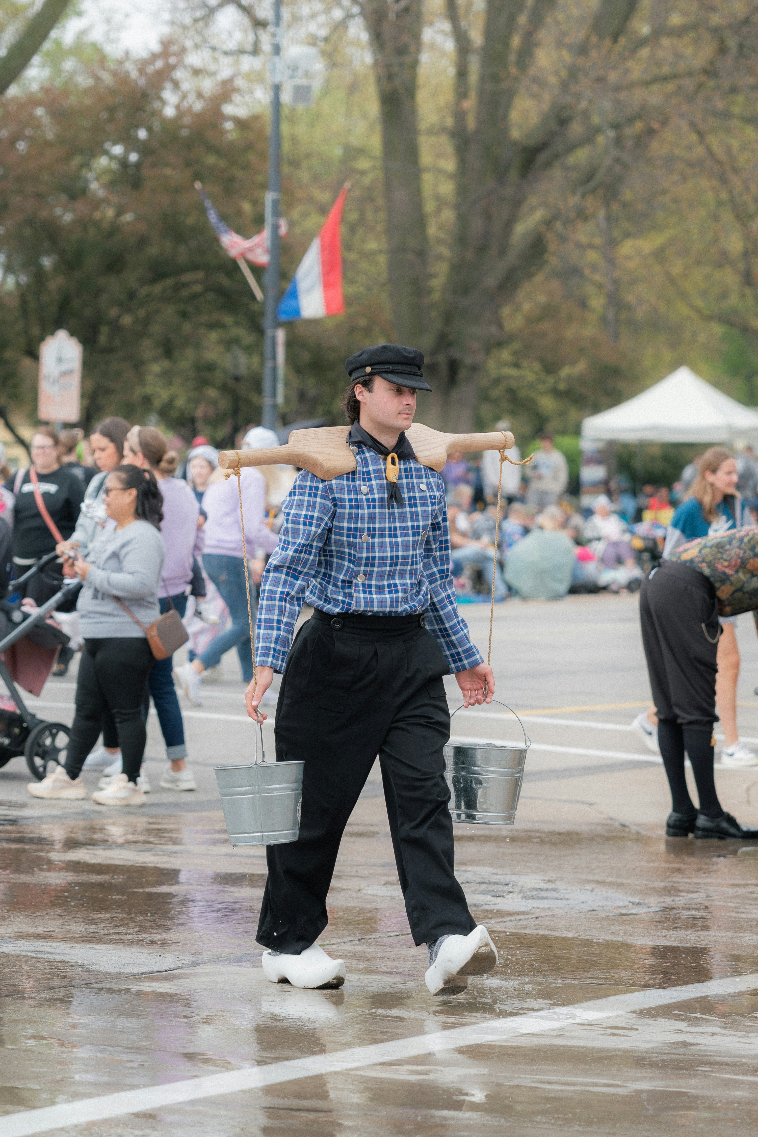 A person dressed in period attire carries two buckets suspended from a yoke during a lively festival, surrounded by a bustling crowd and colorful flags.