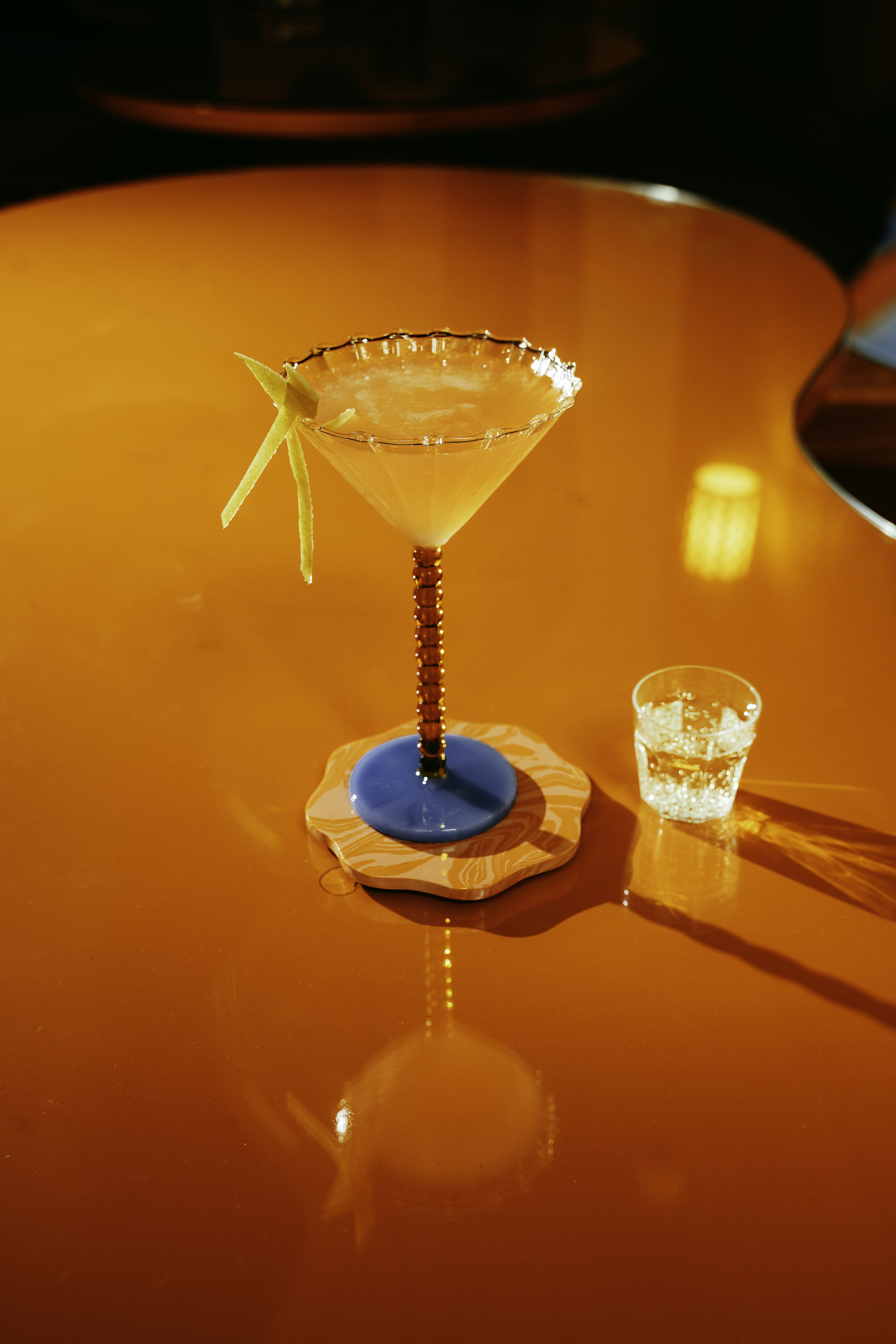 A vibrant cocktail garnished with a lime leaf sits on a colorful coaster beside a small glass of whiskey. The warm glow of ambient lighting enhances the inviting atmosphere.