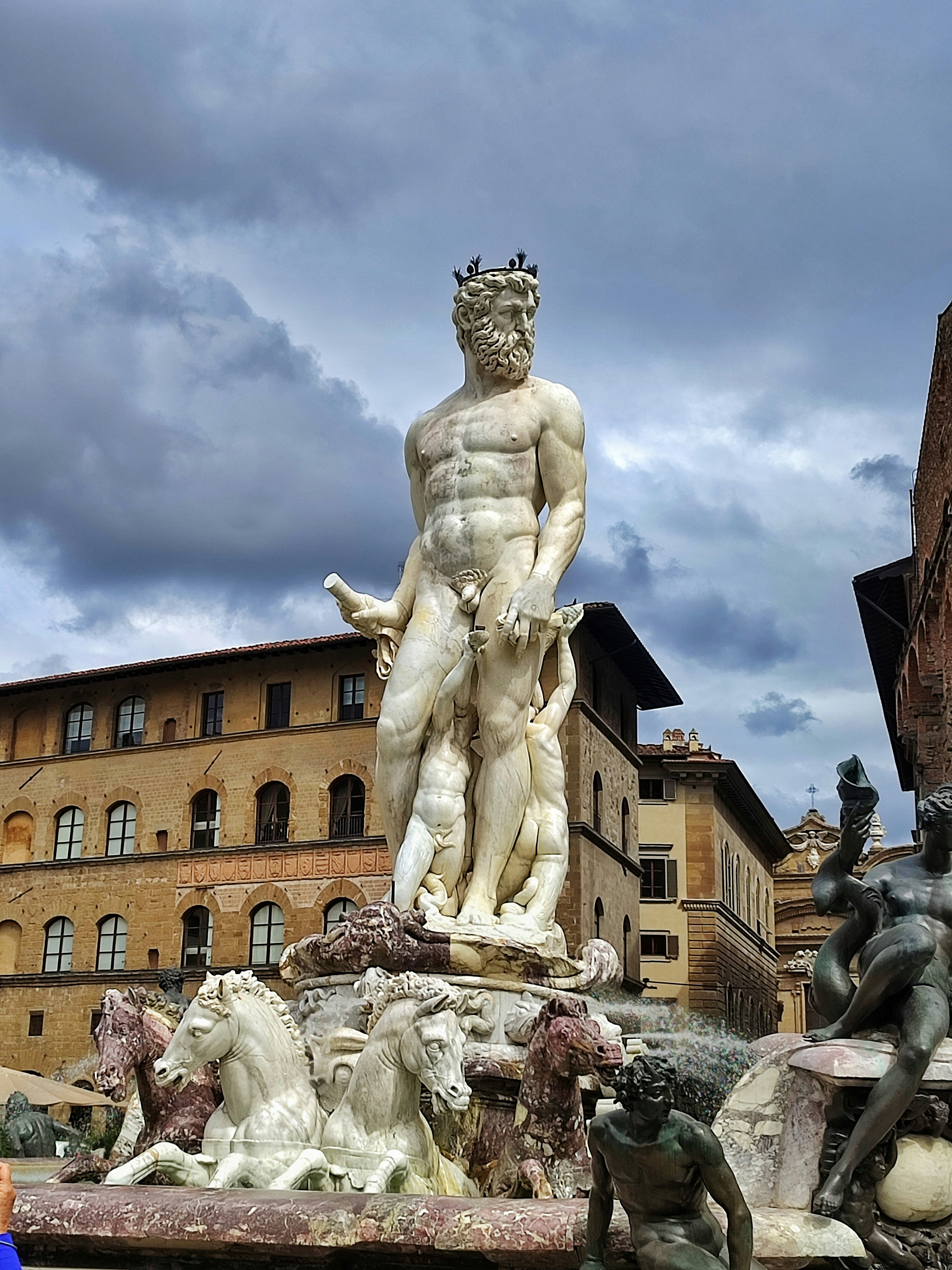 A grand statue depicting a muscular figure surrounded by mythical creatures, set against a dramatic cloudy sky.