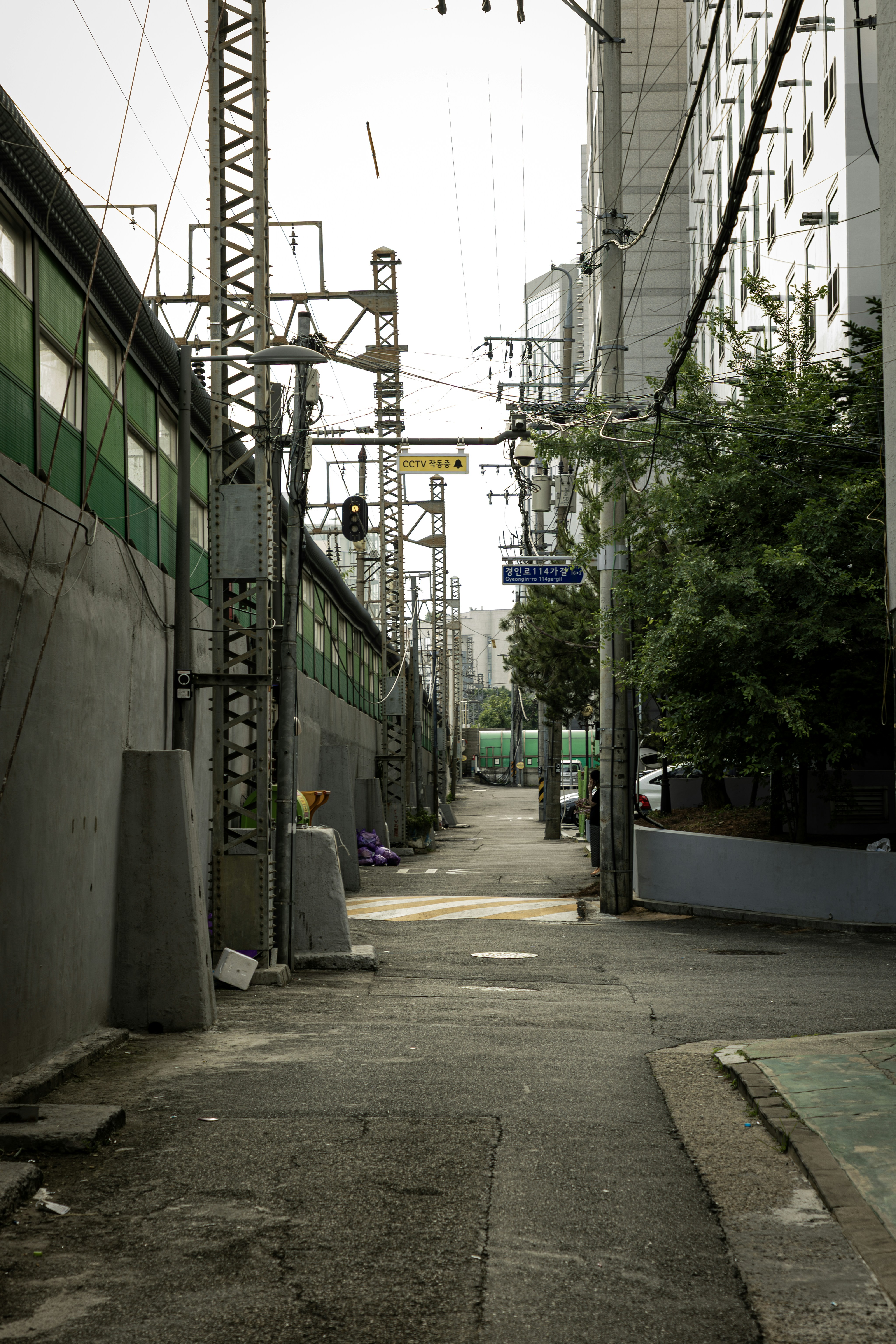 Narrow urban alley with overhead wires, tall buildings, and a green fence, creating a sense of depth and urban exploration.Dohyuk You