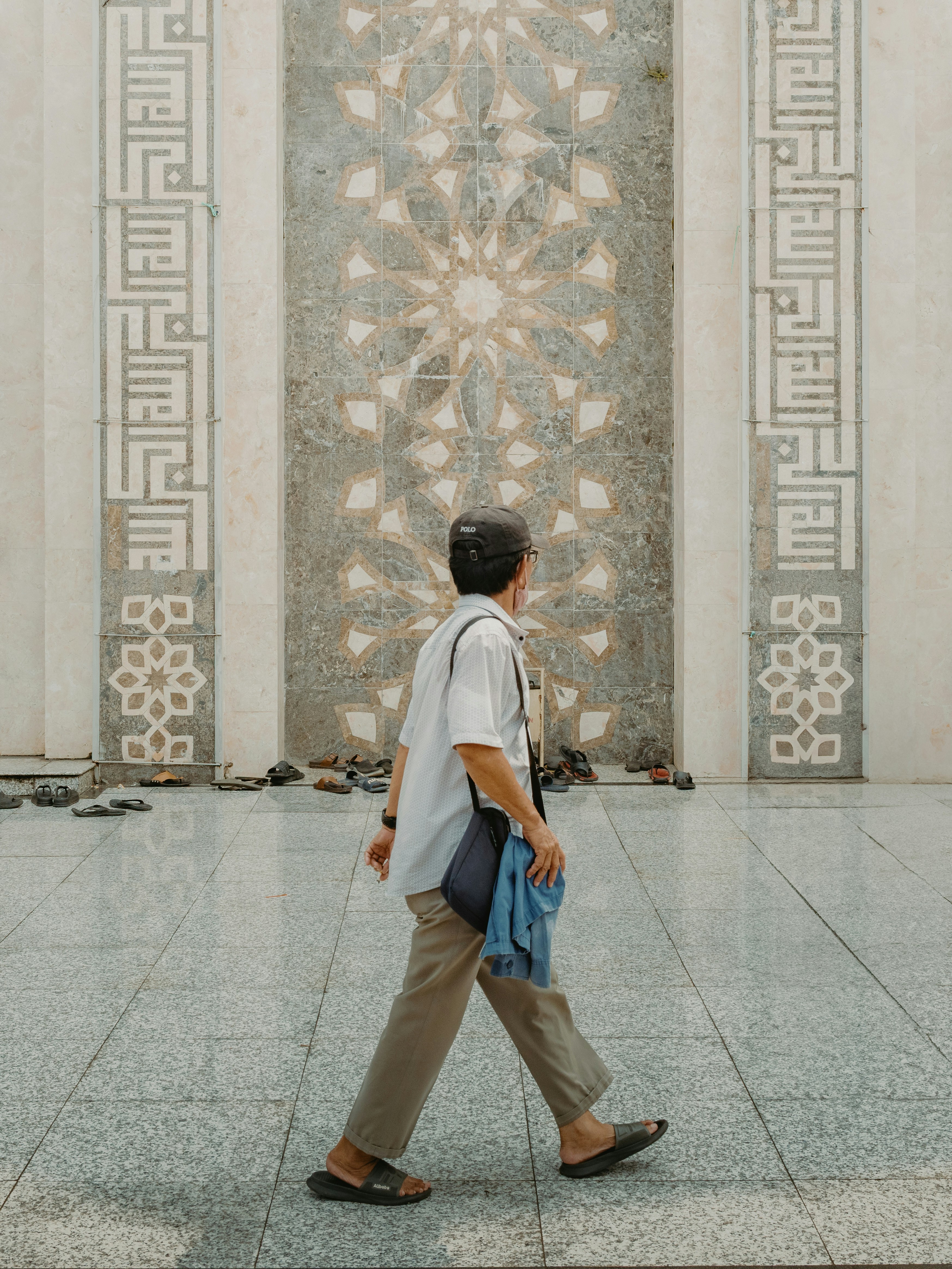 A casually dressed man strides past a grand mosque wall adorned with intricate Islamic patterns under bright daylight.