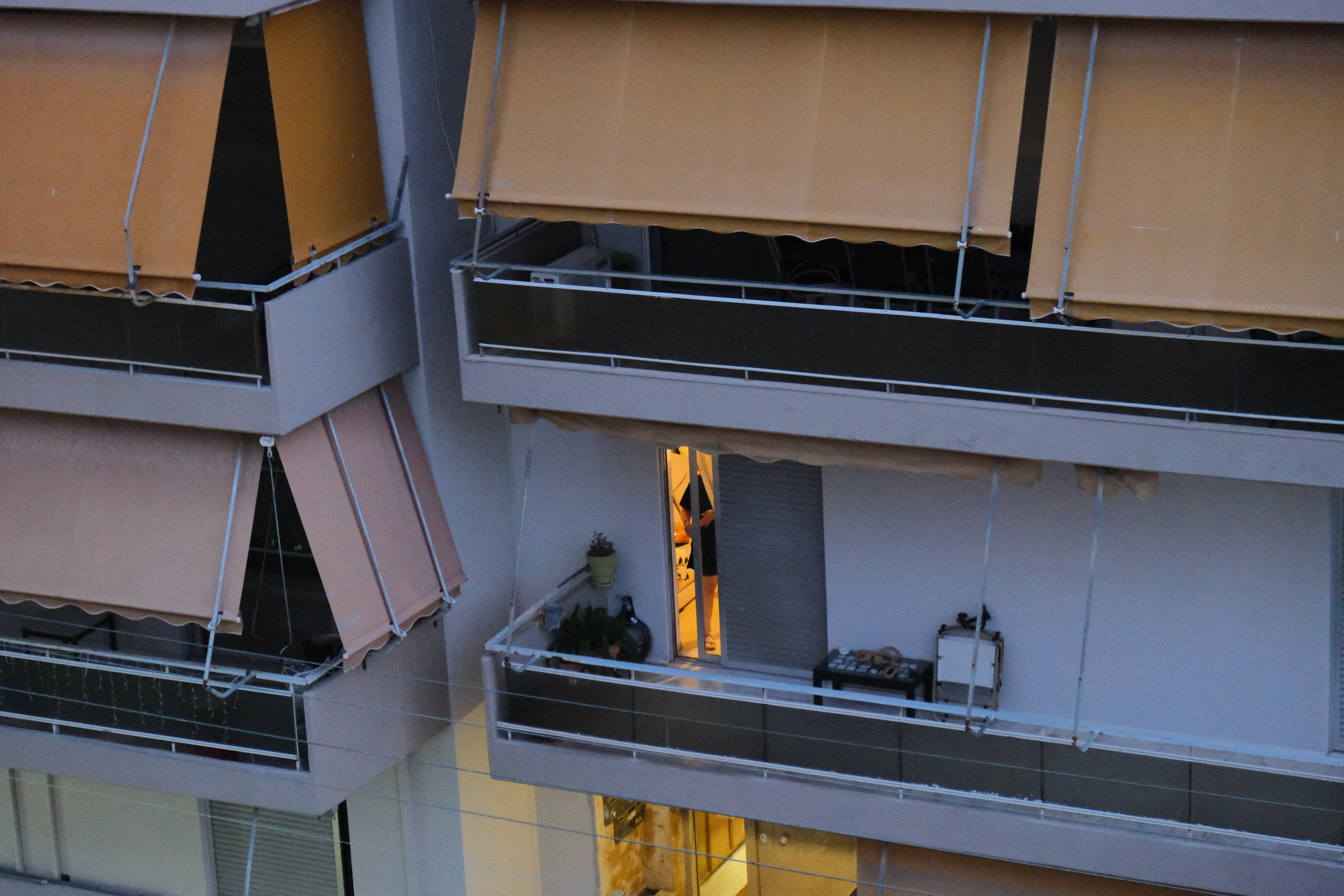 A warmly lit apartment balcony reveals a glimpse of domestic life, framed by the geometric lines of neighboring structures. The scene captures the essence of urban living at dusk.