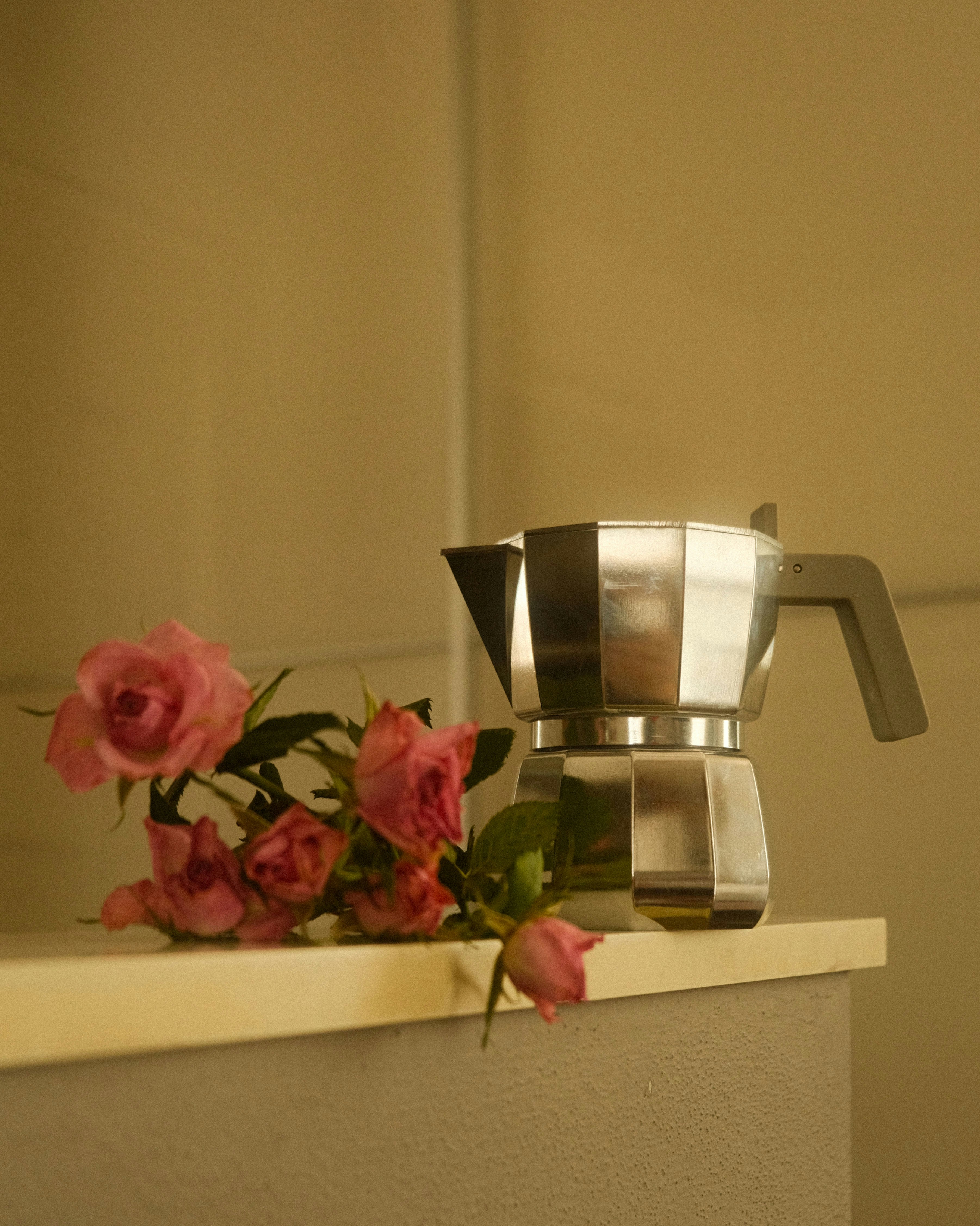 Coffee maker and pink roses rest on a ledge.
