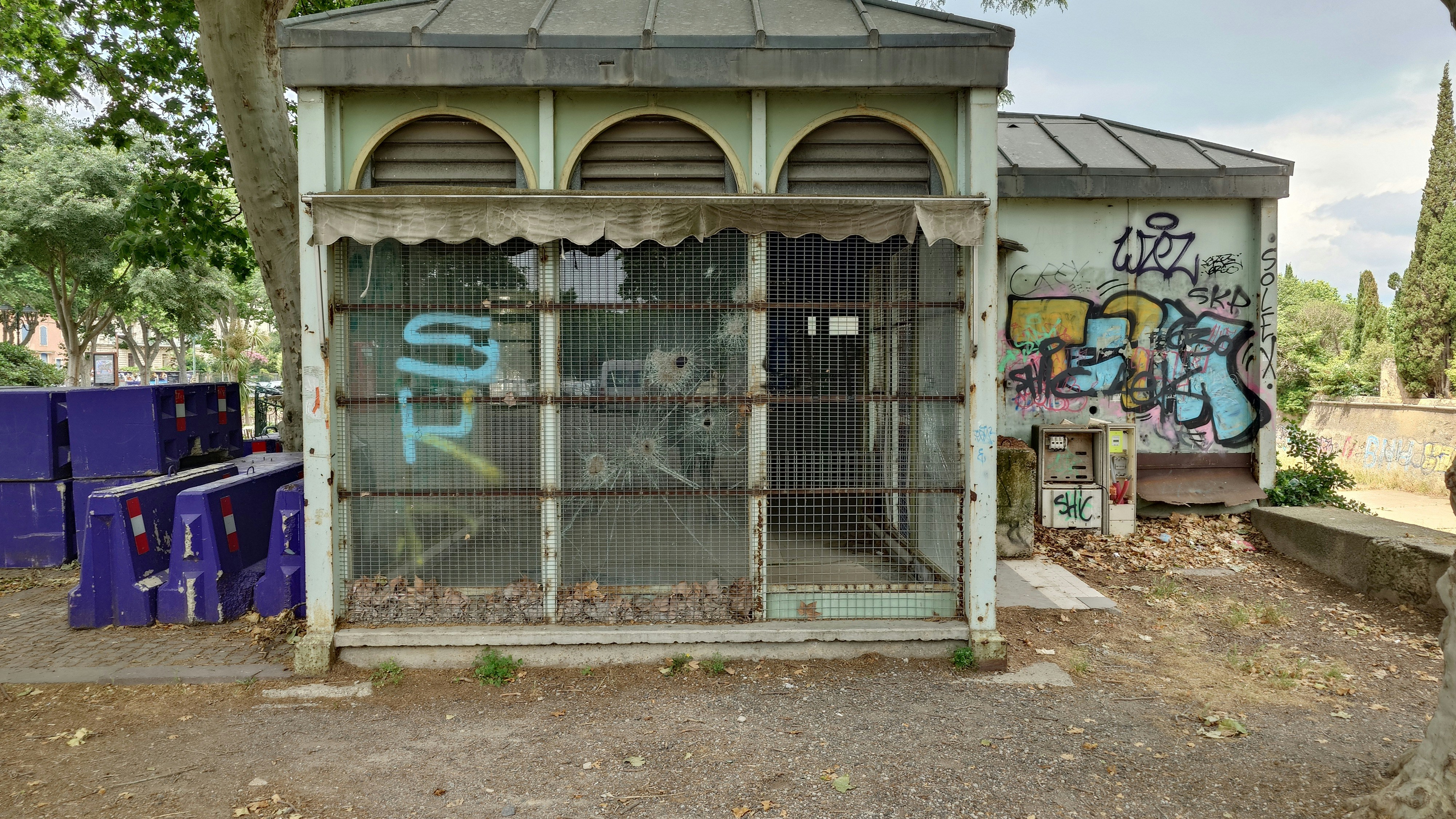 Abandoned structure with a mesh enclosure, adorned with graffiti, set in an urban landscape. Surrounding elements hint at a once-vibrant area now faded.