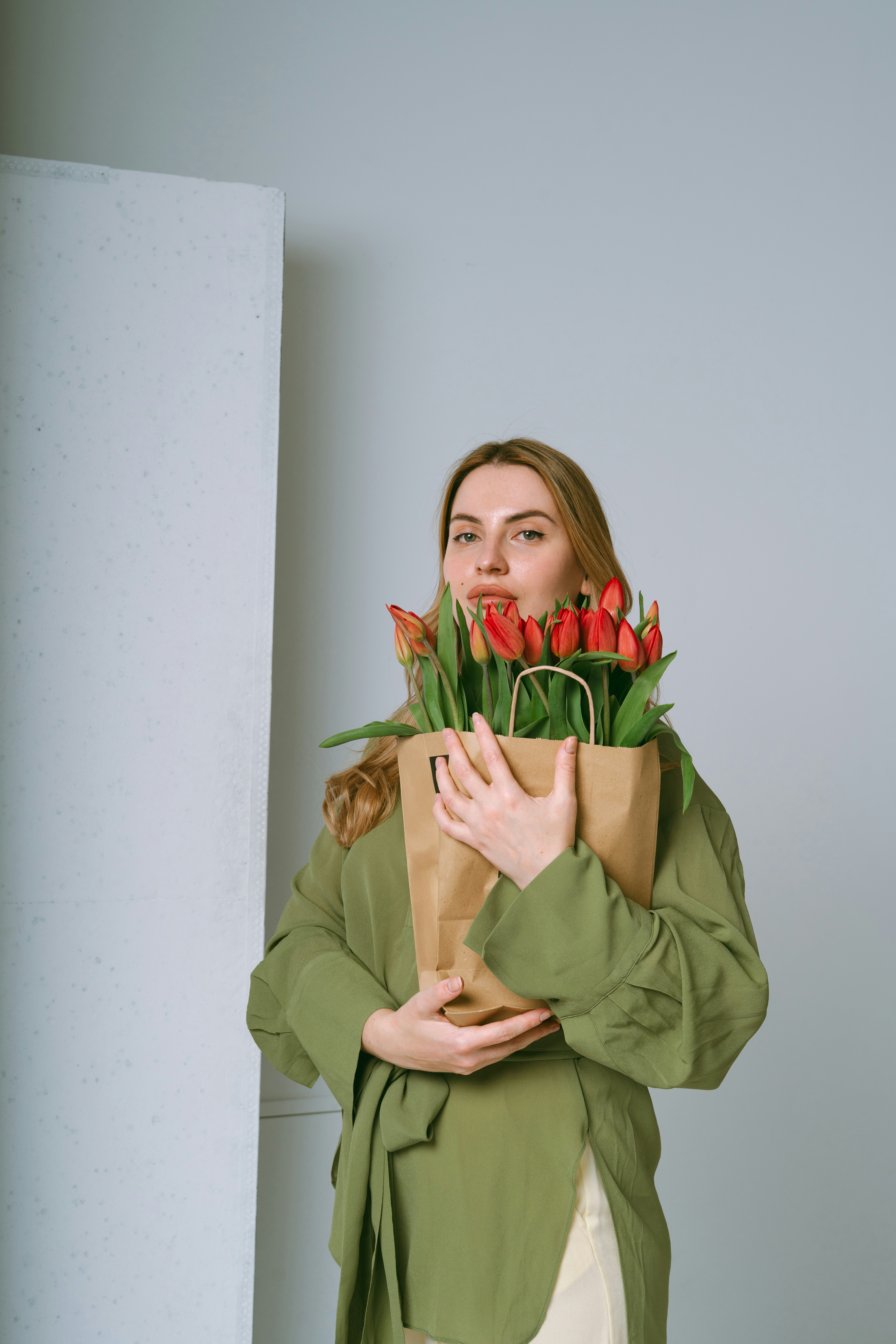 Woman in a green blouse holding a bouquet of red tulips in a paper bag against a soft gray background.