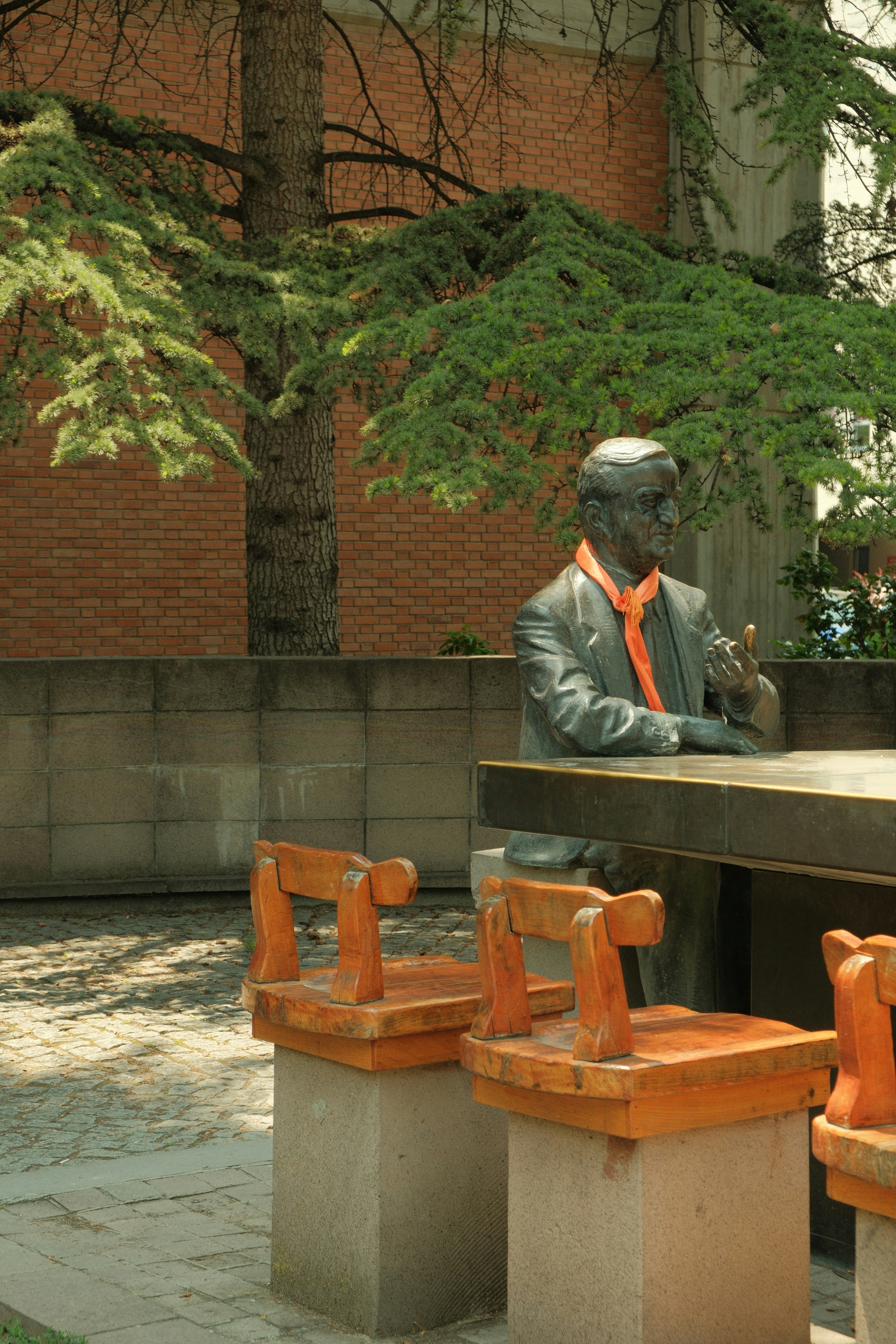 Bronze statue of a seated philosopher wearing an orange scarf, positioned at a table with empty wooden chairs around it. The setting features natural greenery and brick walls.