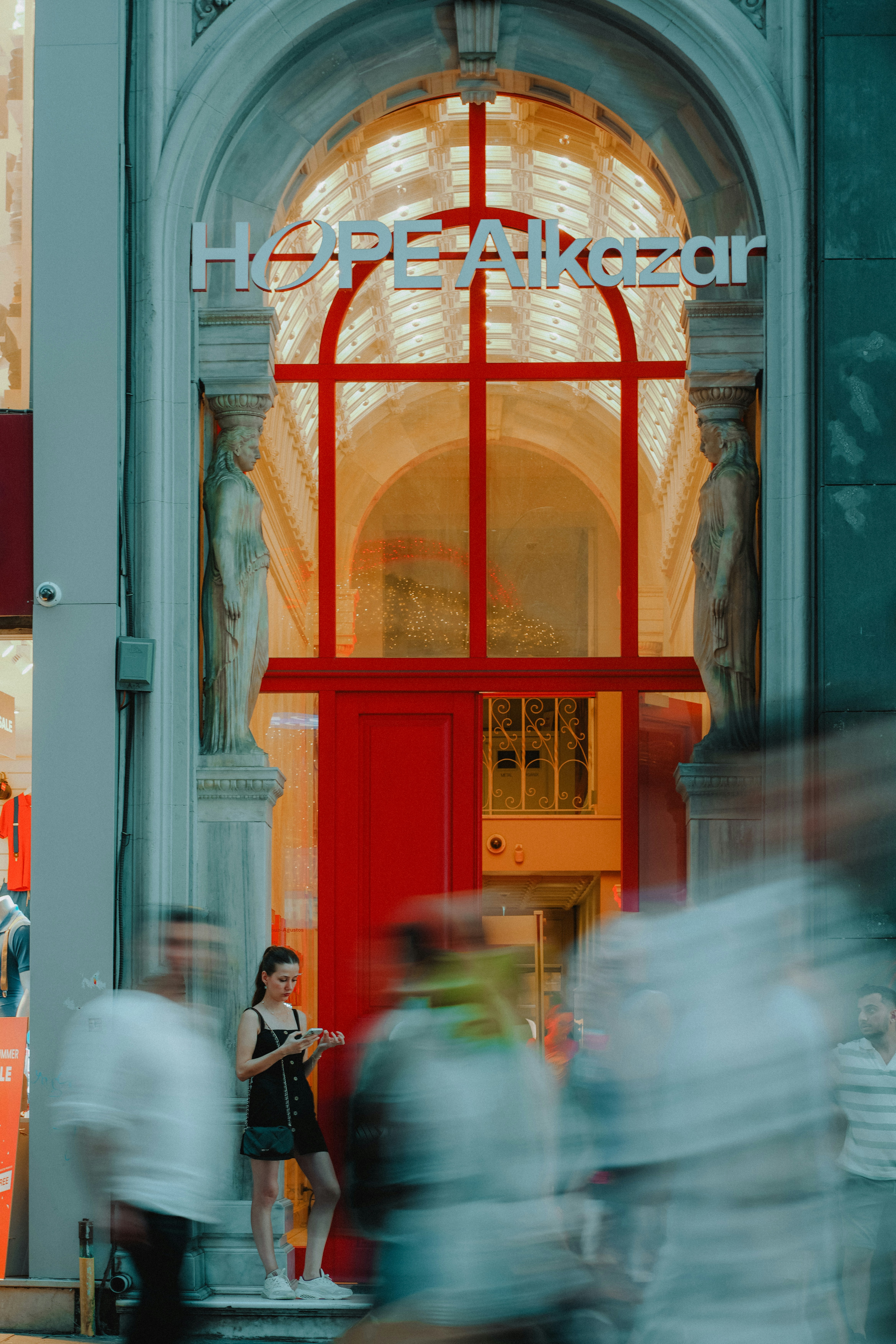 A woman stands calmly by a vibrant red entrance, surrounded by a blur of pedestrians in motion. The contrasting stillness highlights the hustle of city life.