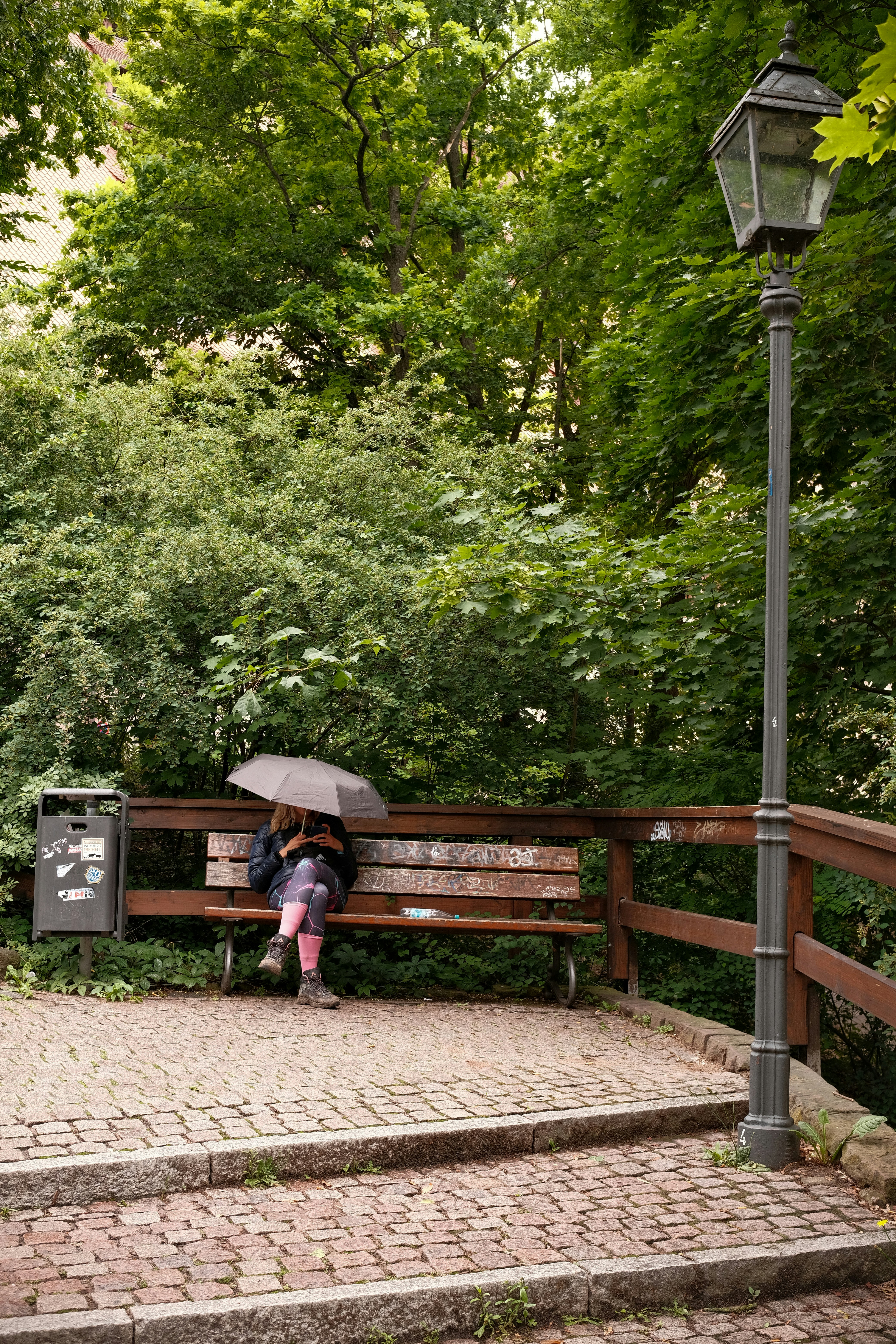 A person seated on a bench under an umbrella, surrounded by lush greenery, enjoying a moment of solitude in an urban park.