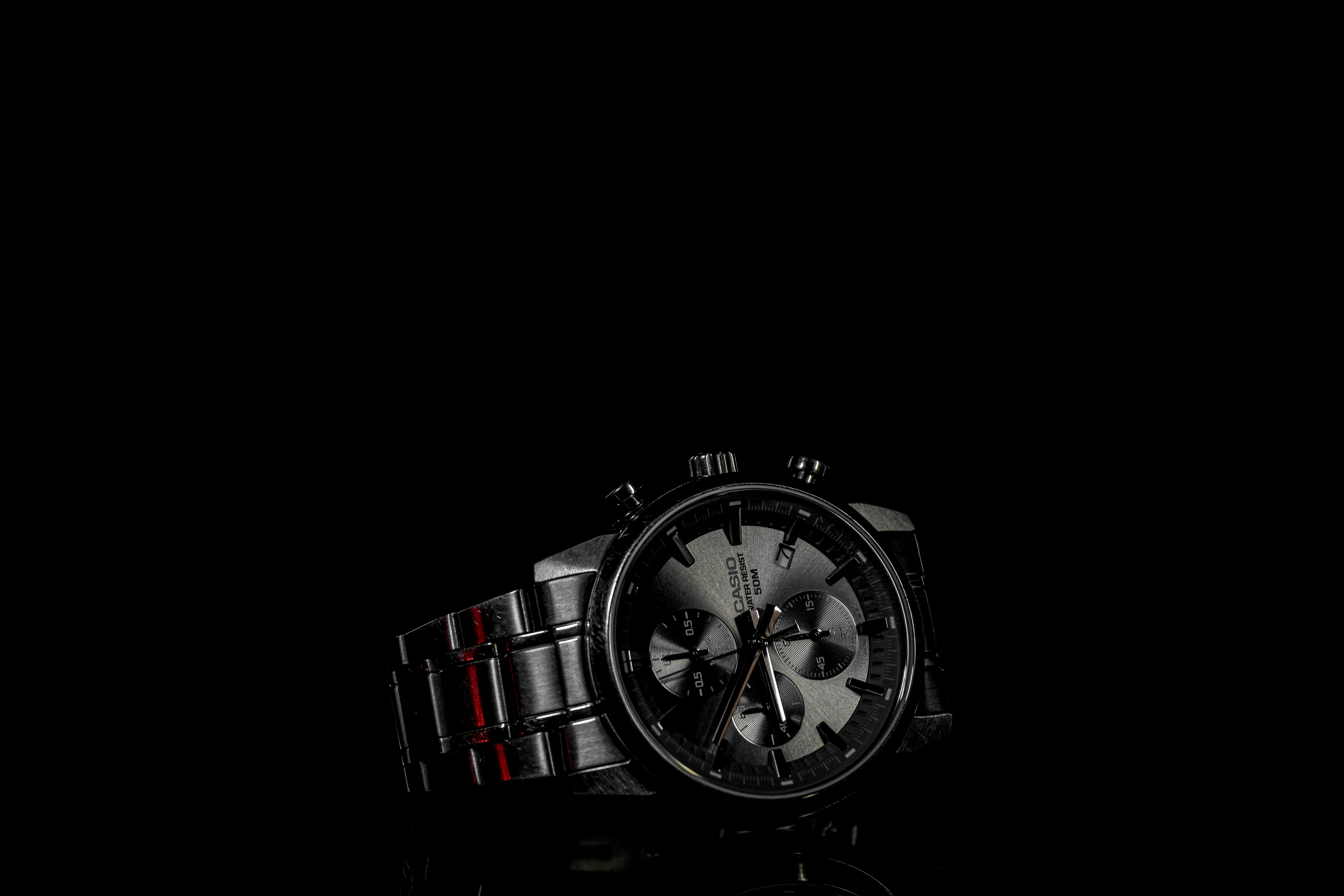 A sleek black chronograph watch with a silver bracelet, elegantly illuminated against a dark backdrop, highlighting its intricate details.