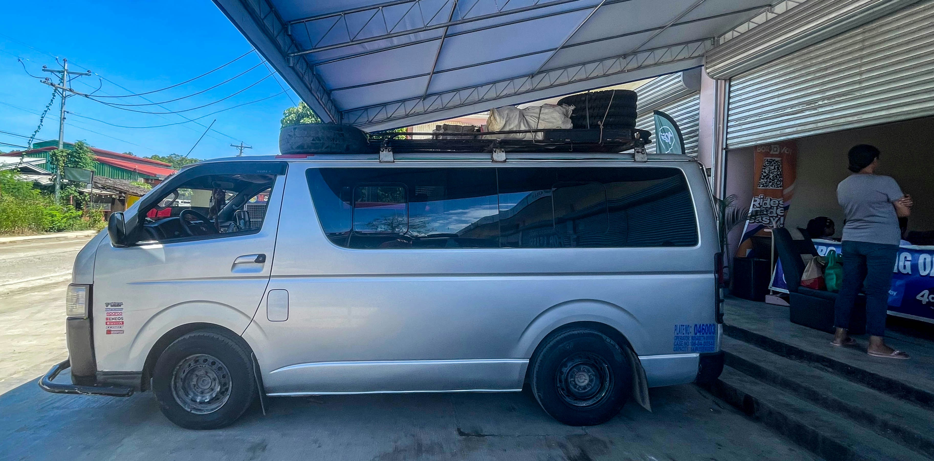 Silver van parked under a canopy, showcasing urban transportation. A few people are visible in the background, hinting at daily life in the area.