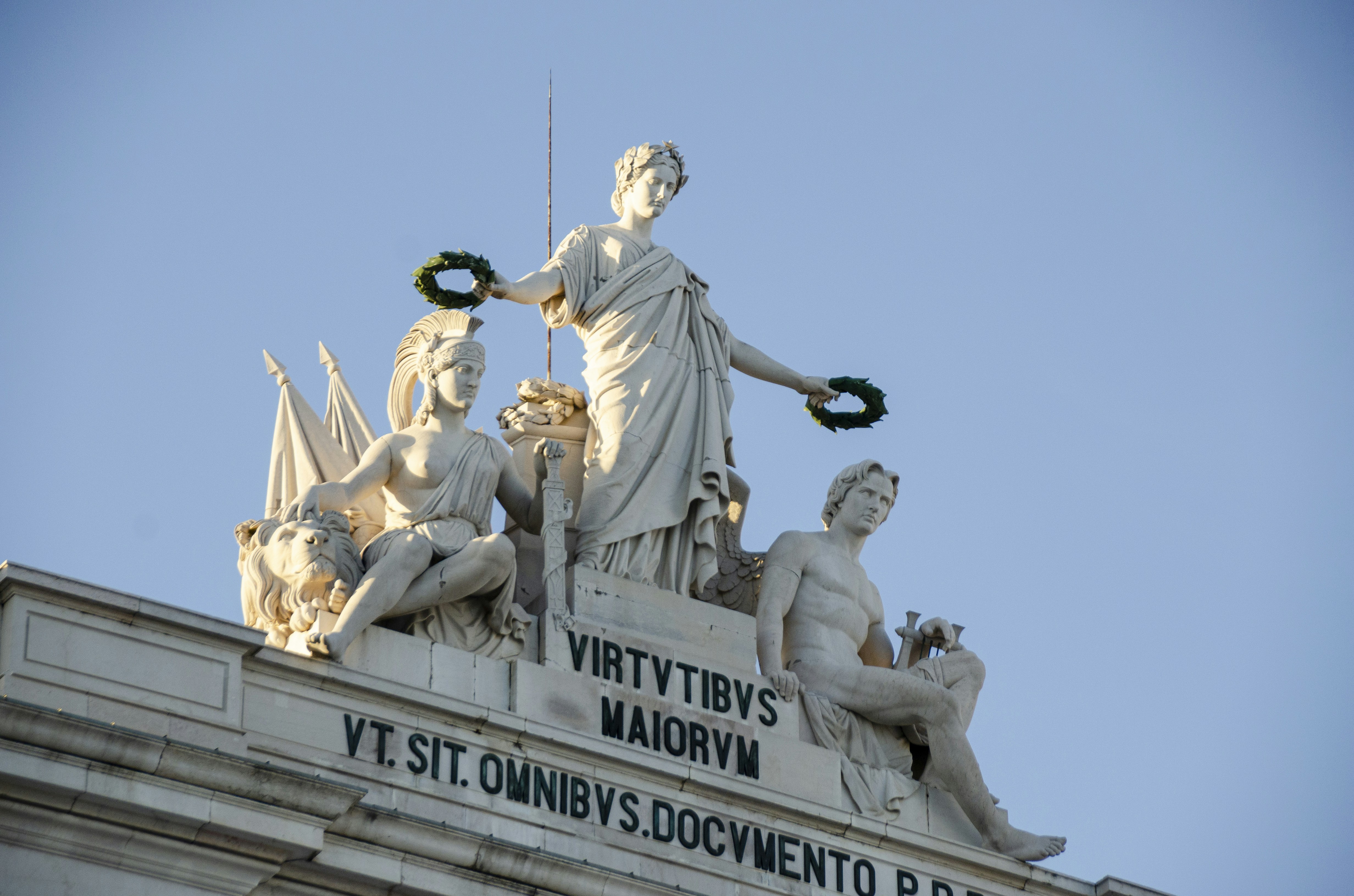 Classical sculptures representing virtue, adorned with wreaths, atop a grand architectural structure against a clear sky.