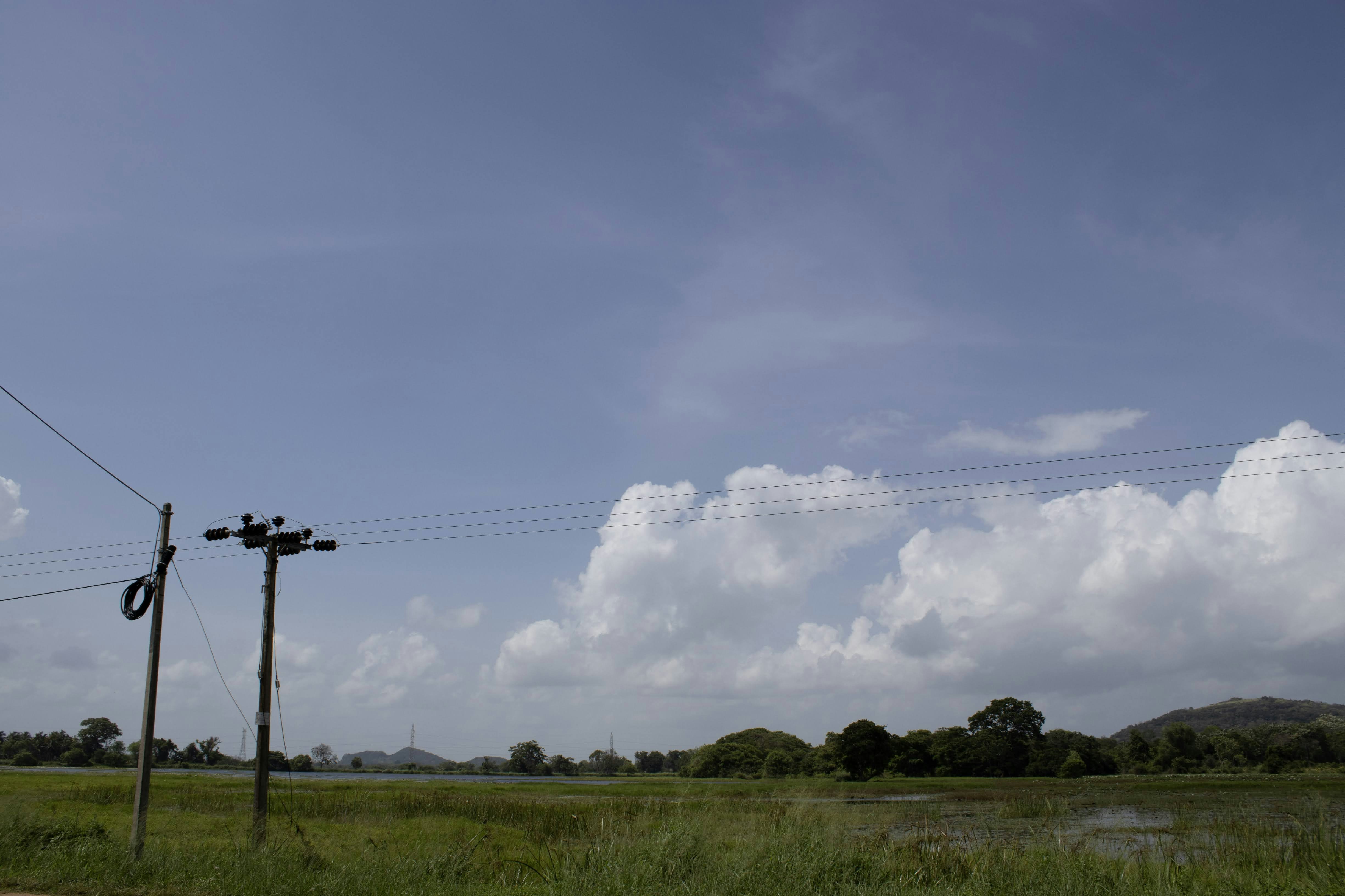 A serene landscape featuring power lines and lush greenery under a vast sky filled with clouds.